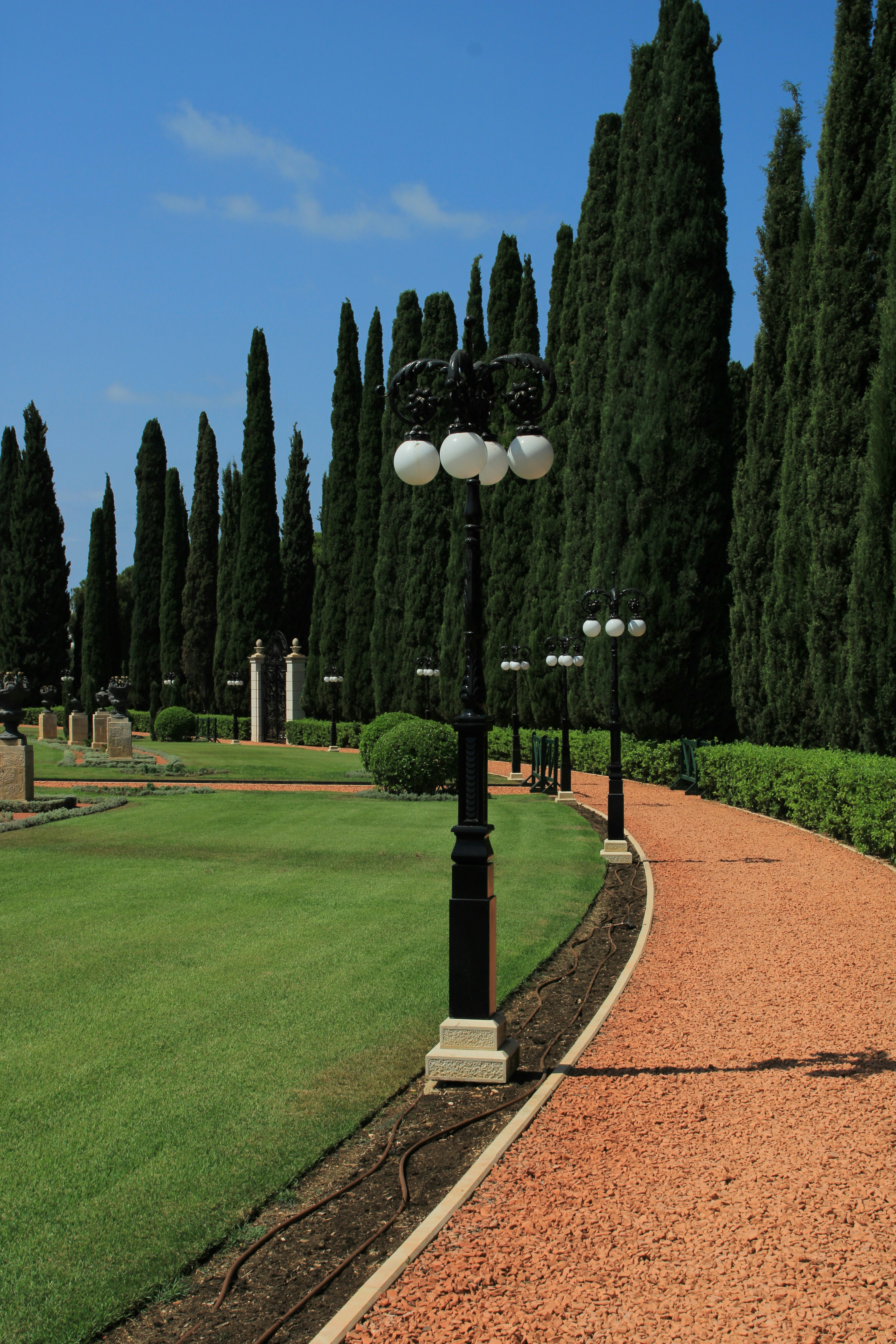 A sunlit vertical view of a curved red gravel path lined with ornate black lamp posts and white globe lights, bordered by a pristine green lawn and a wall of tall cypress trees in the peaceful Bahai Gardens of Akko, Israel