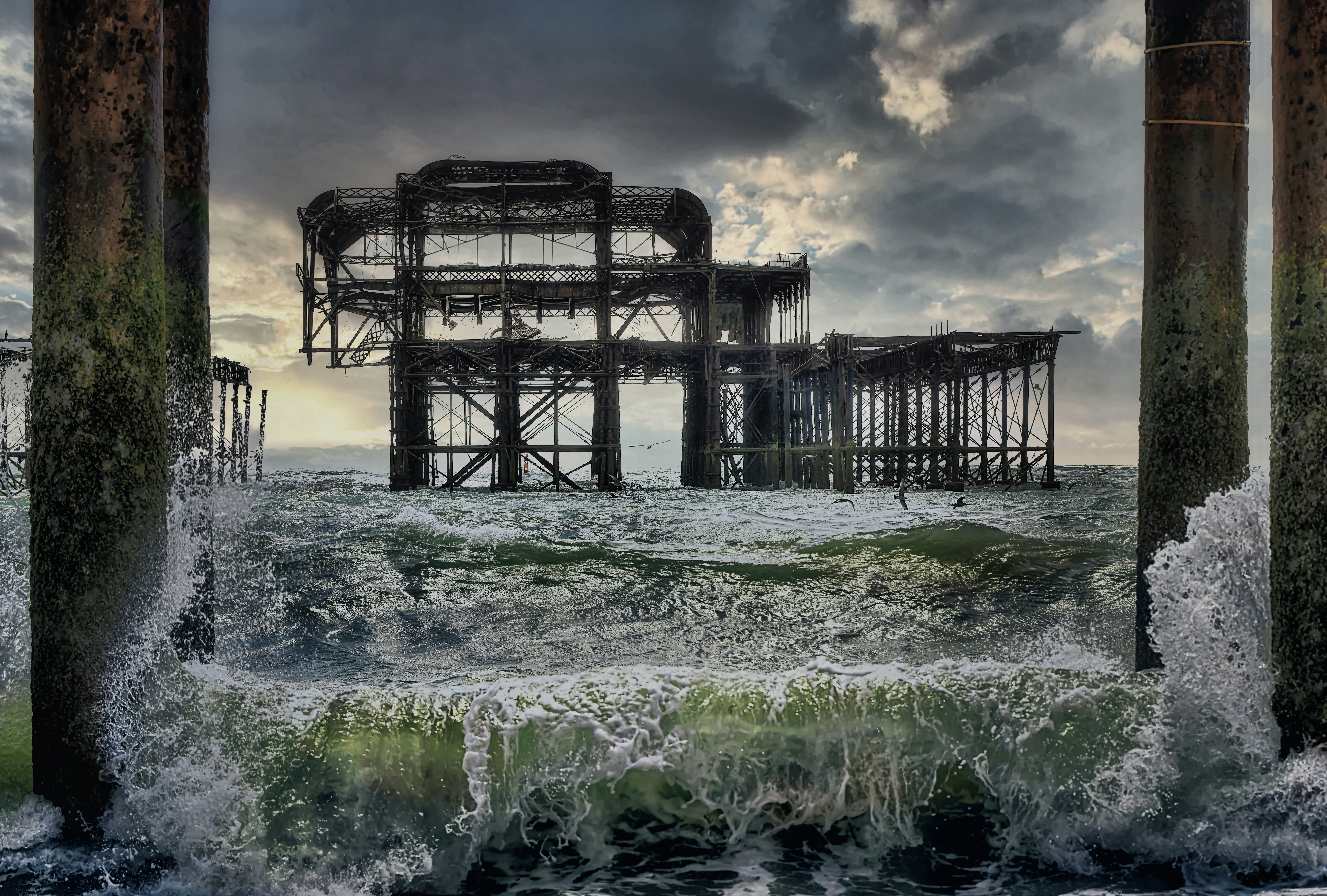 Weathered skeletal structure of a pier stands against a turbulent sea, framed by breaking waves and dark clouds; a moody, industrial ruin captured at sea level with dramatic contrast.