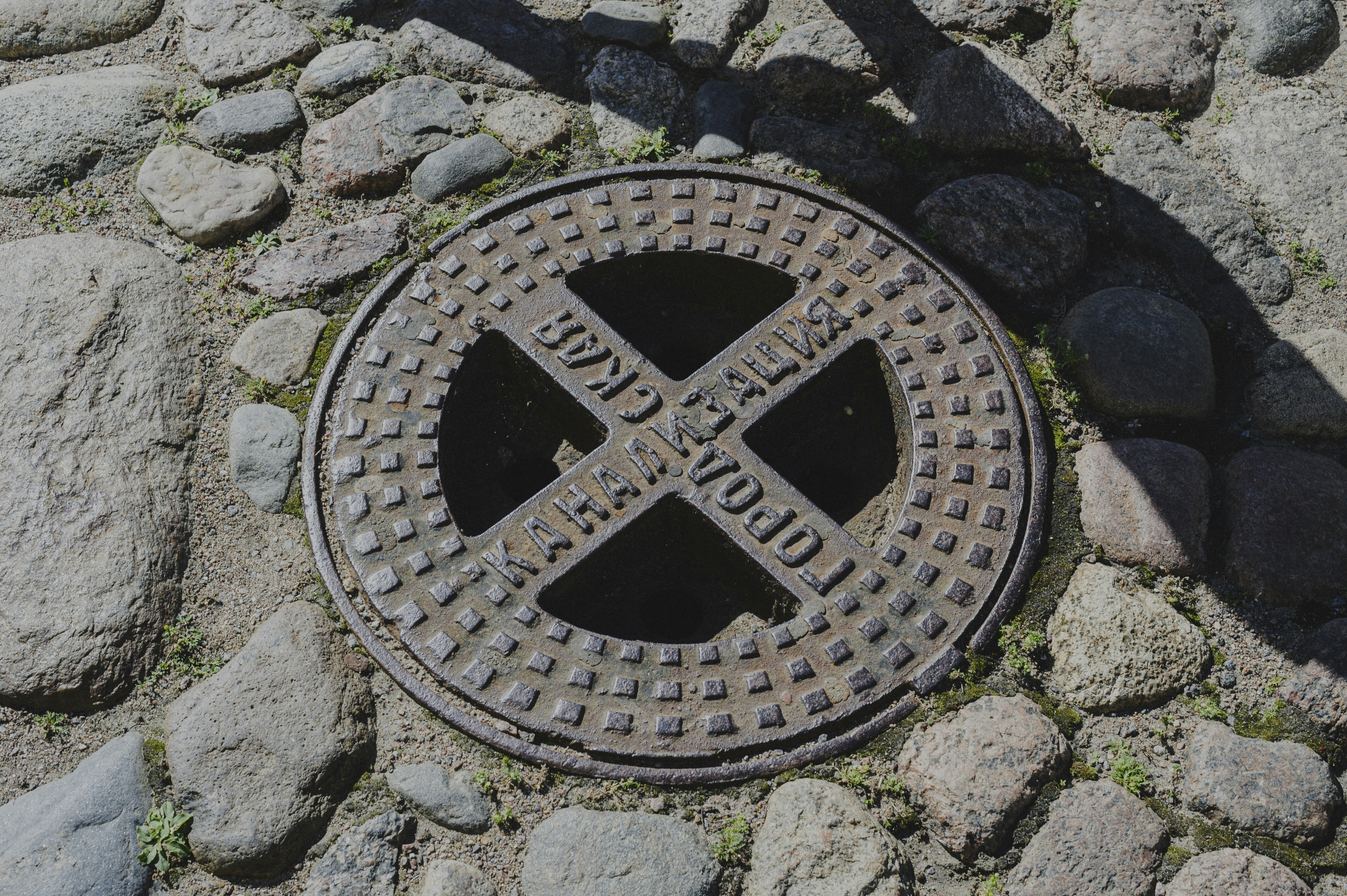 Crosshatched Circle: A Metal Drain Cover Embedded in Pebbles