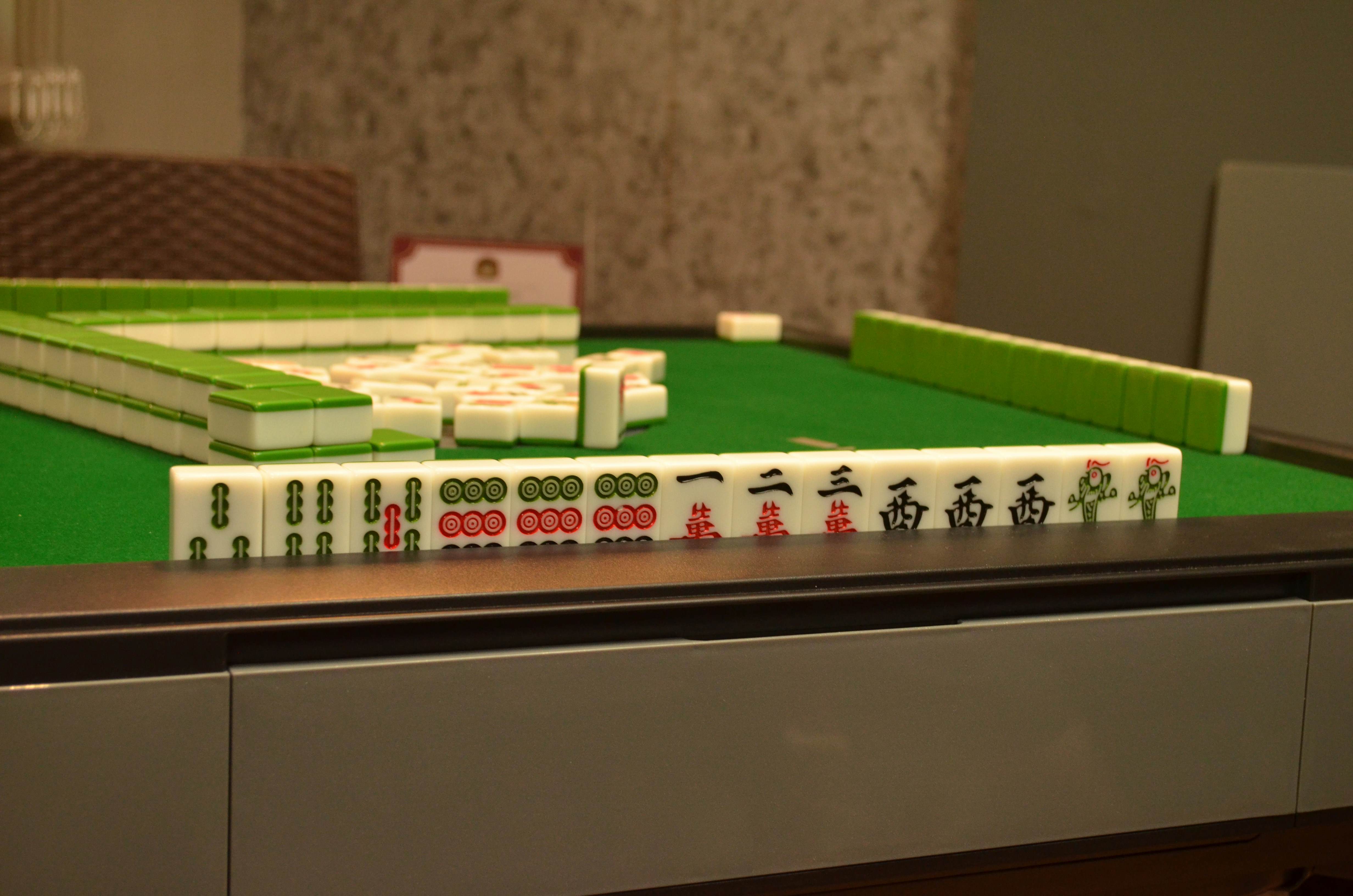 Close-up view of a mahjong table with tiles arranged for a game, showing suits and dots; a calm, indoor setting with a green felt surface and a partial wall behind. (Logo/panel not evident).