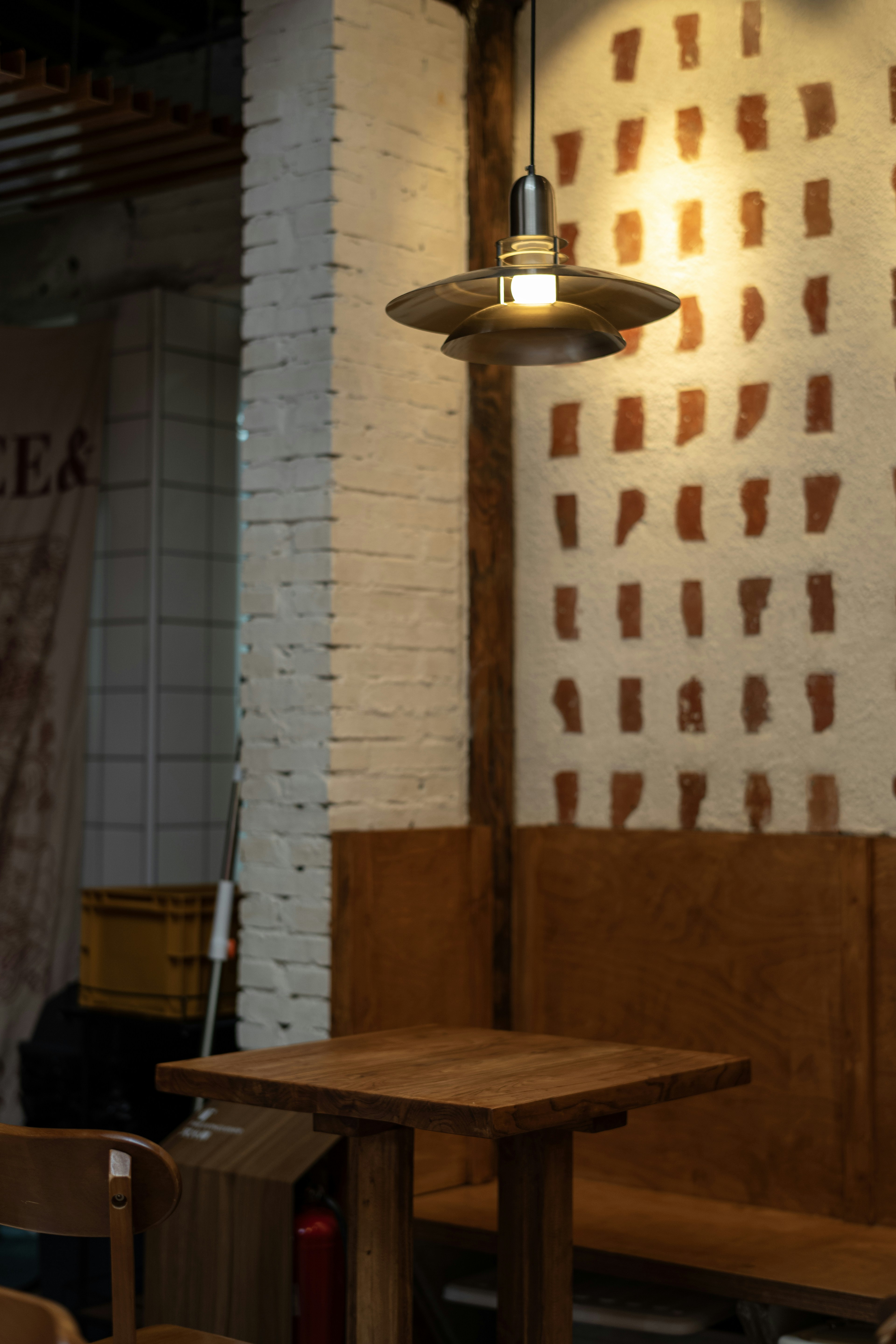 A warmly lit cafe corner with a hanging industrial lamp above a wooden table and bench, against a textured wall with vertical brick accents.
