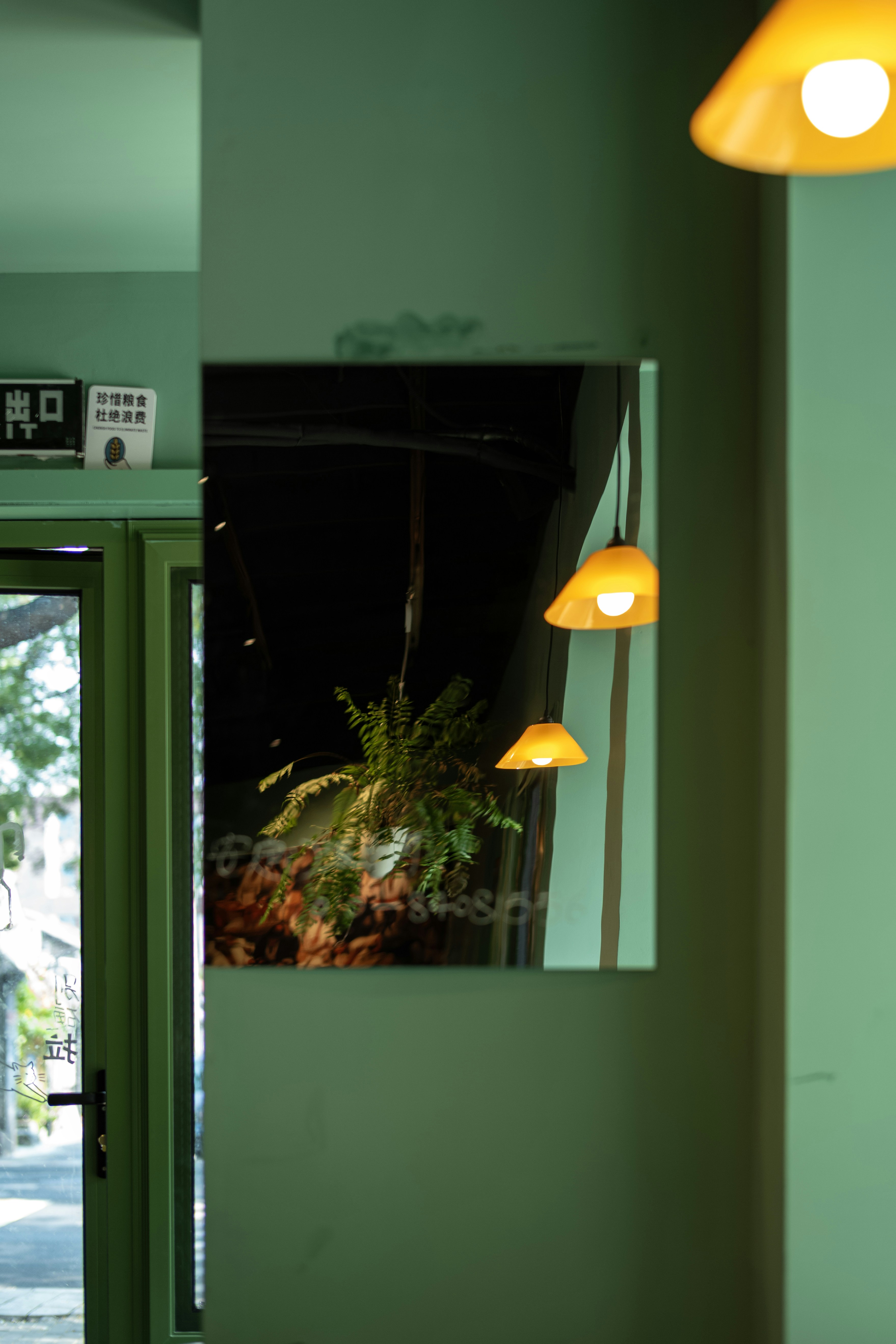 A vertical interior scene featuring green walls, a doorway, and a line of yellow pendant lamps casting warm light; a plant sits within a cut-out window, hinting at a cafe or studio space.