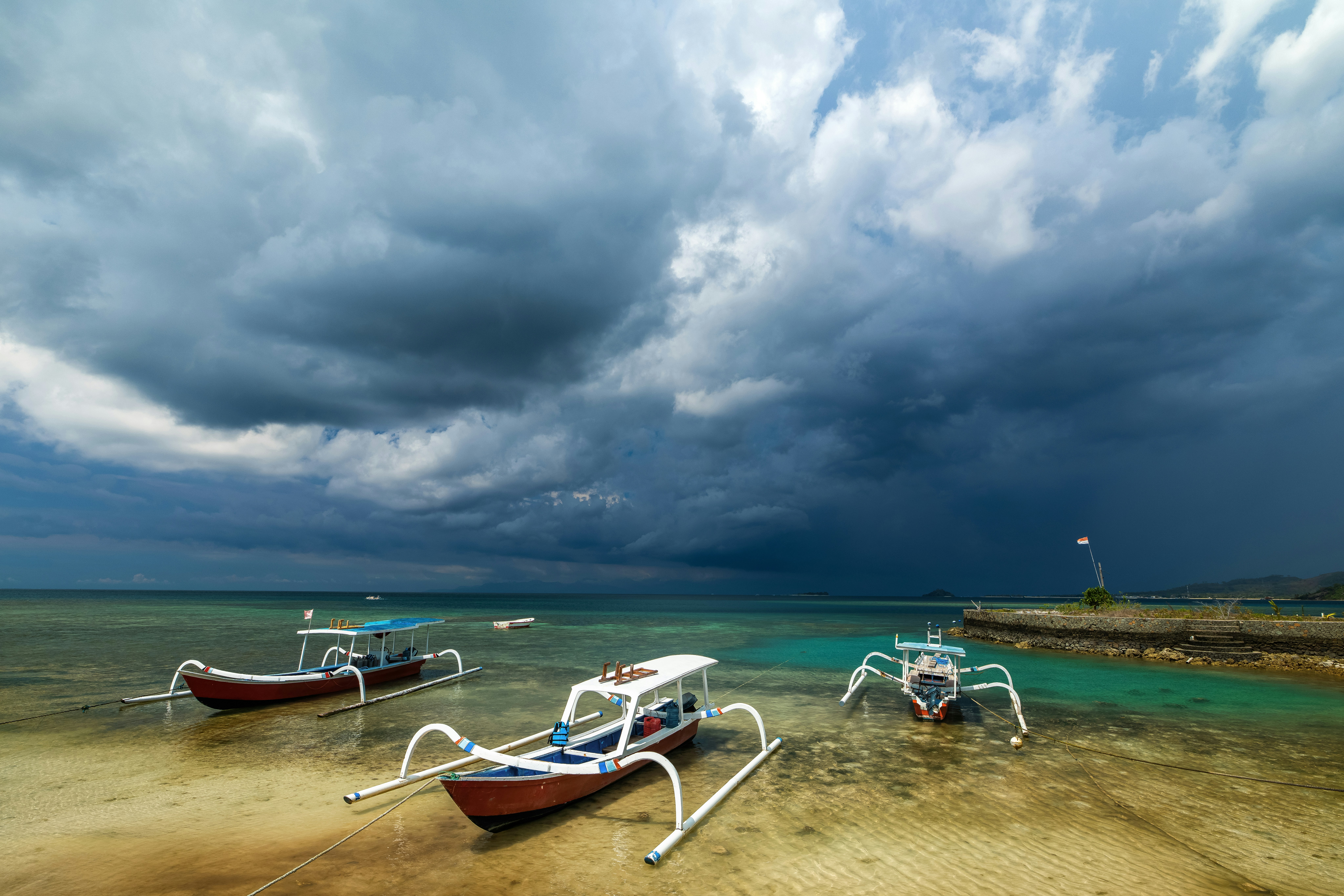 Colorful boats sit on a shallow tropical shoreline as dark storm clouds gather over turquoise water, hinting at an approaching squall.