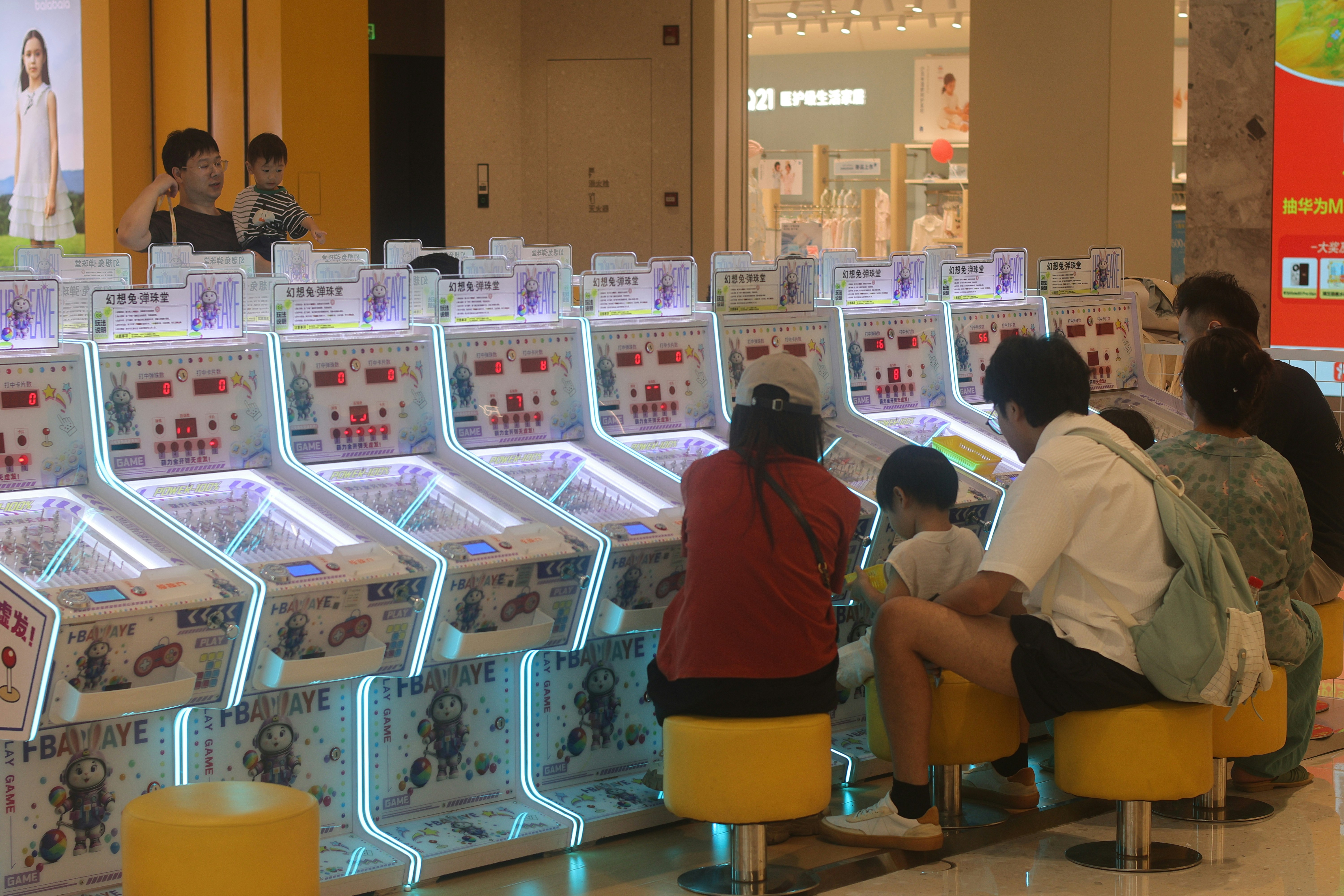 Row of illuminated prize machines in a shopping mall, with customers seated on yellow stools facing the cabinets; a busy, colorful indoor scene with people engaged in the activity.