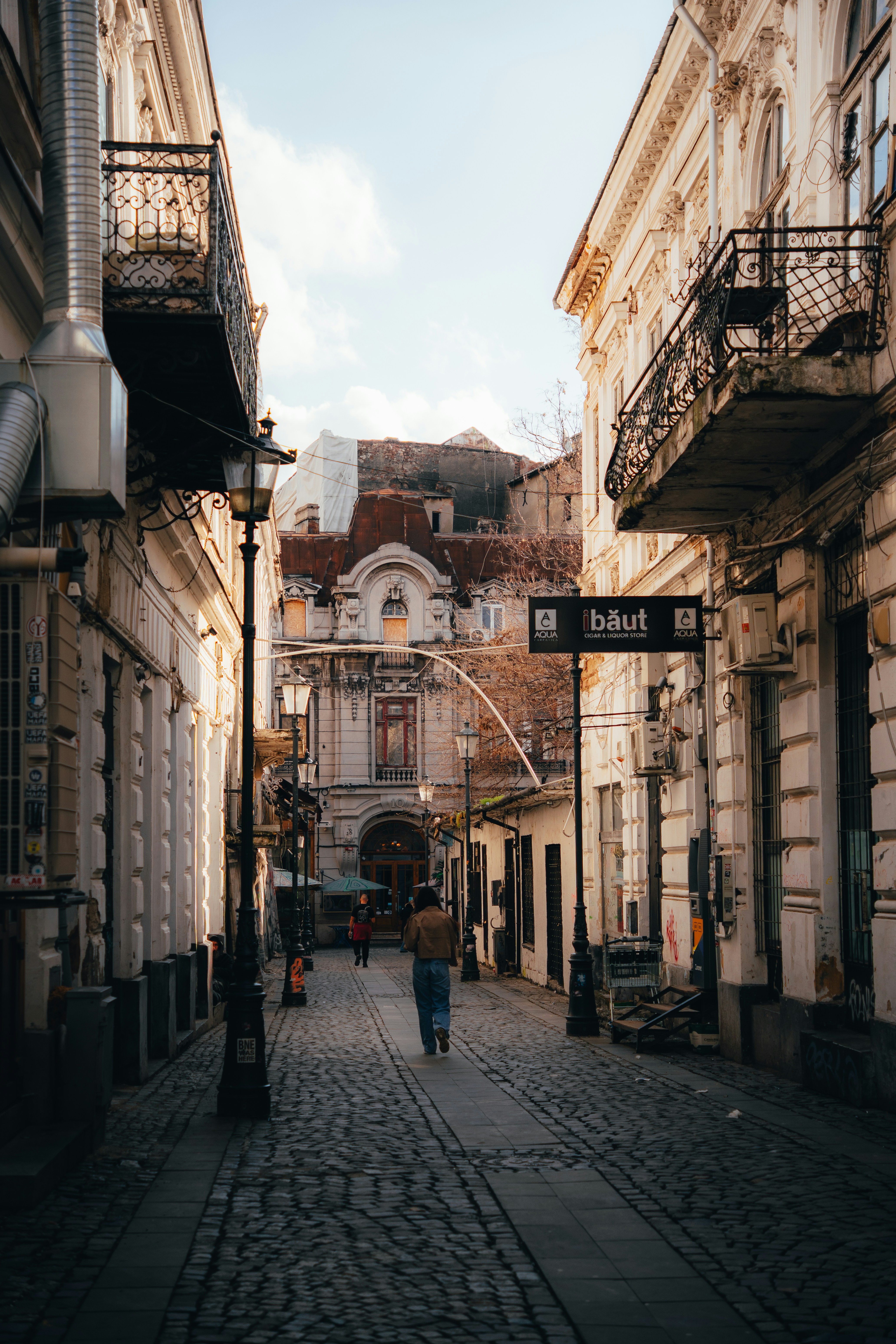 Cobblestone street lined with classic European buildings leading to a grand arched facade; pedestrians stroll under warm daylight in a quaint urban alley. (Logo-like sign visible on right.)