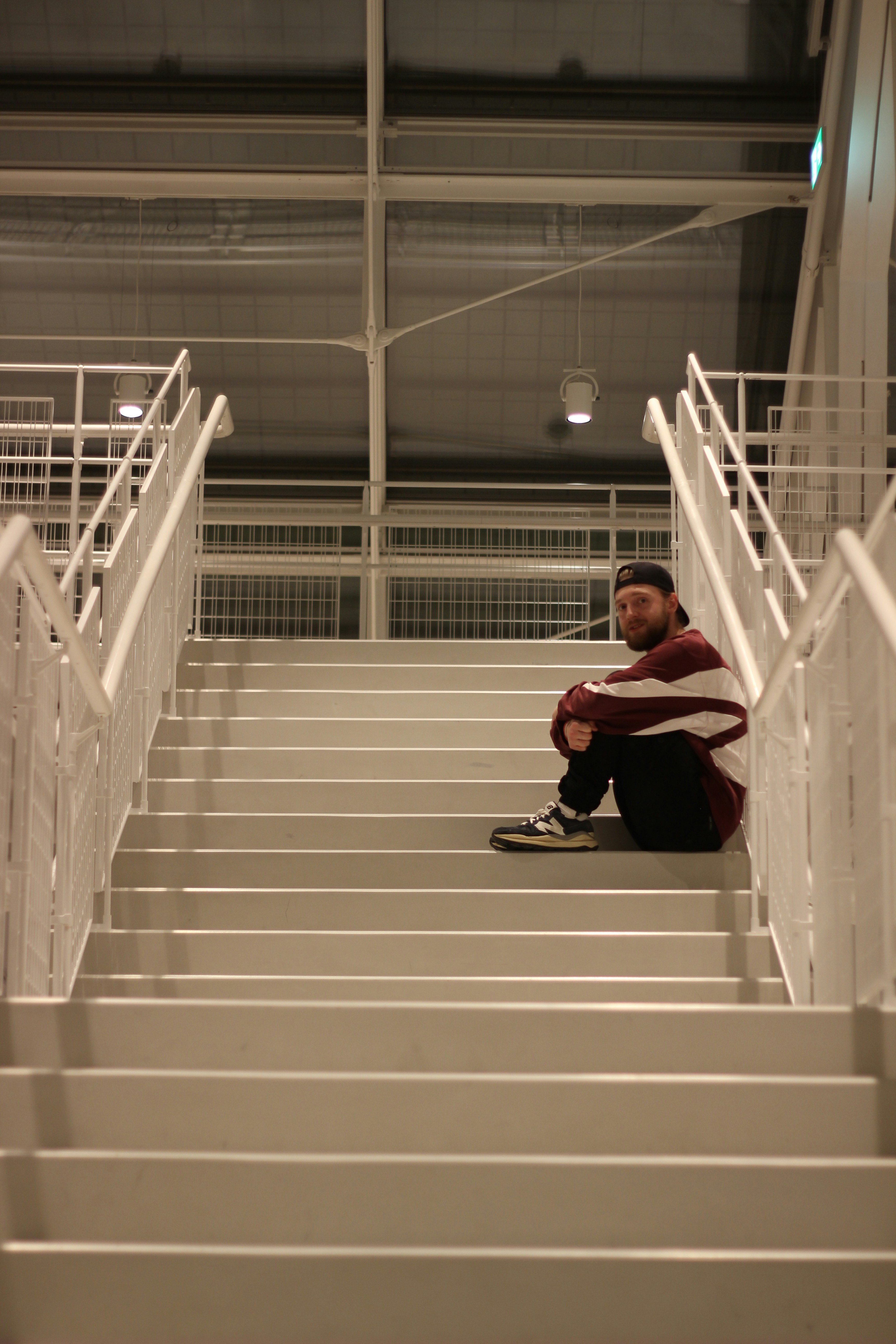 A solitary man sits on modern white stairs in an industrial setting, framed by metal railings and overhead lighting. The scene emphasizes solitude within a spacious, architectural interior.