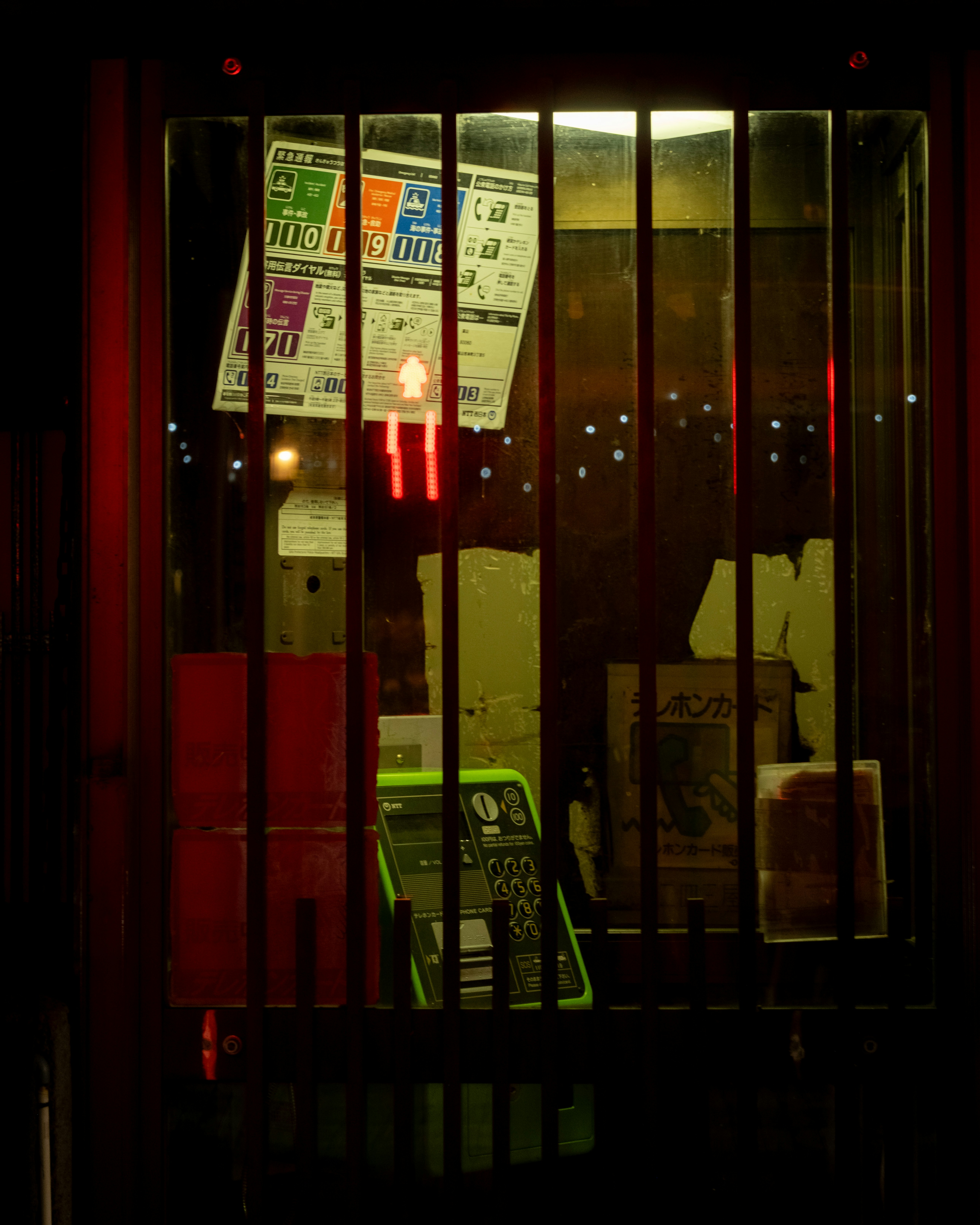 A green public payphone inside a dark glass booth with vertical metal bars