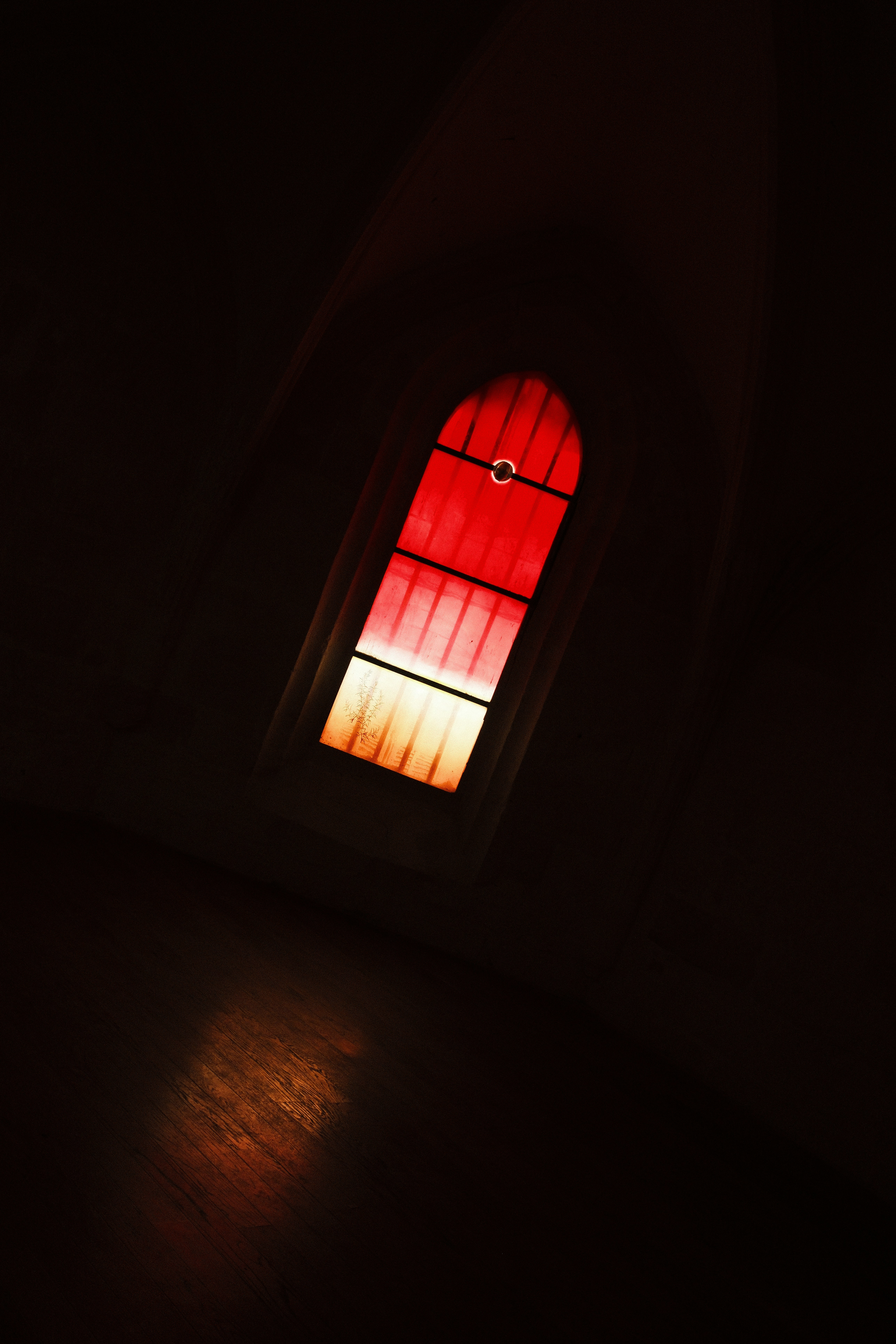Red stained-glass window emits warm glow in an almost empty, shadow-filled interior. Vertical arch and light spill create a moody, contemplative scene.