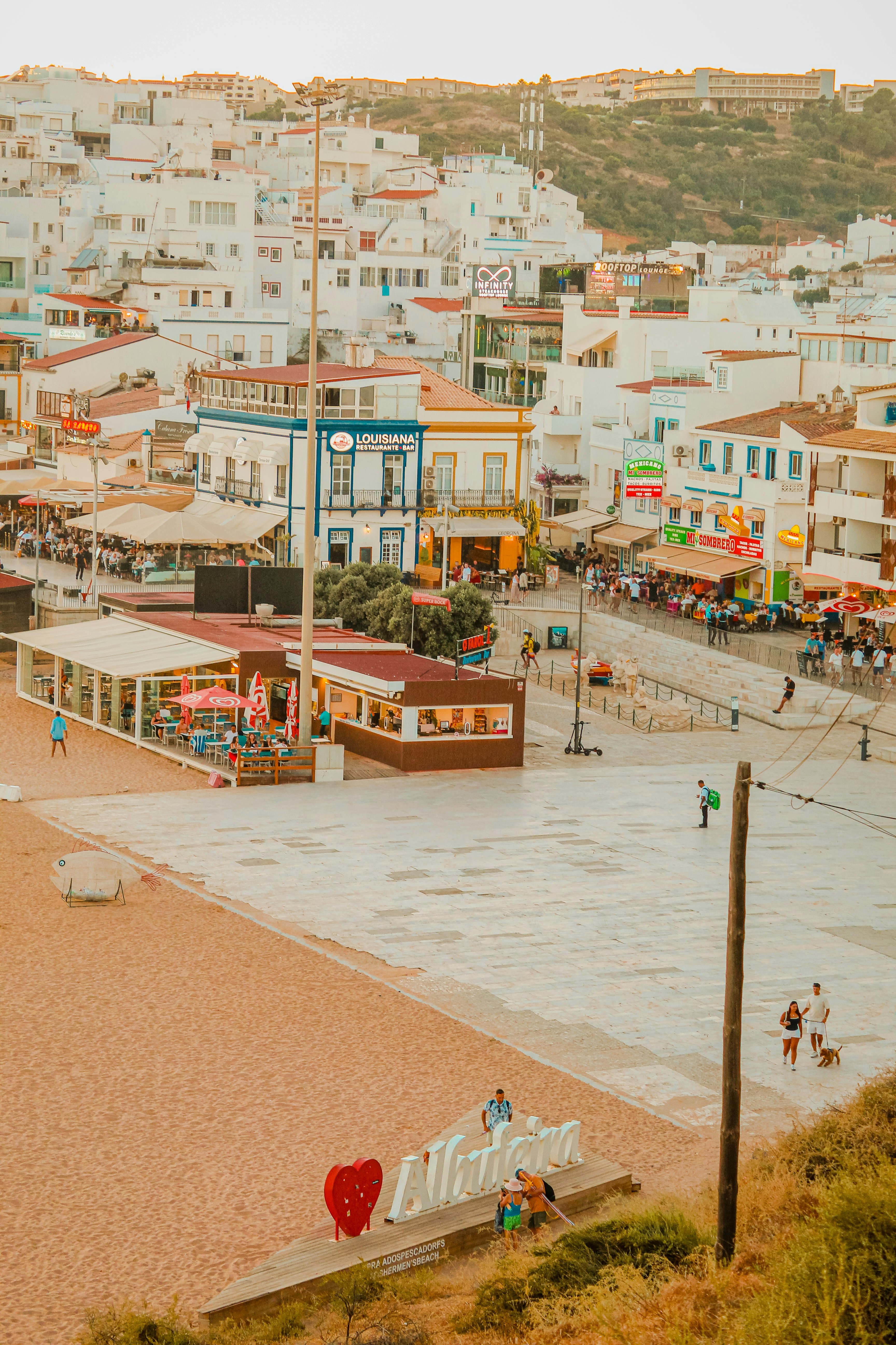 Sunlit Coastal Town Square: A Mosaic of Shops, Beachfront Paths, and Distant Hills