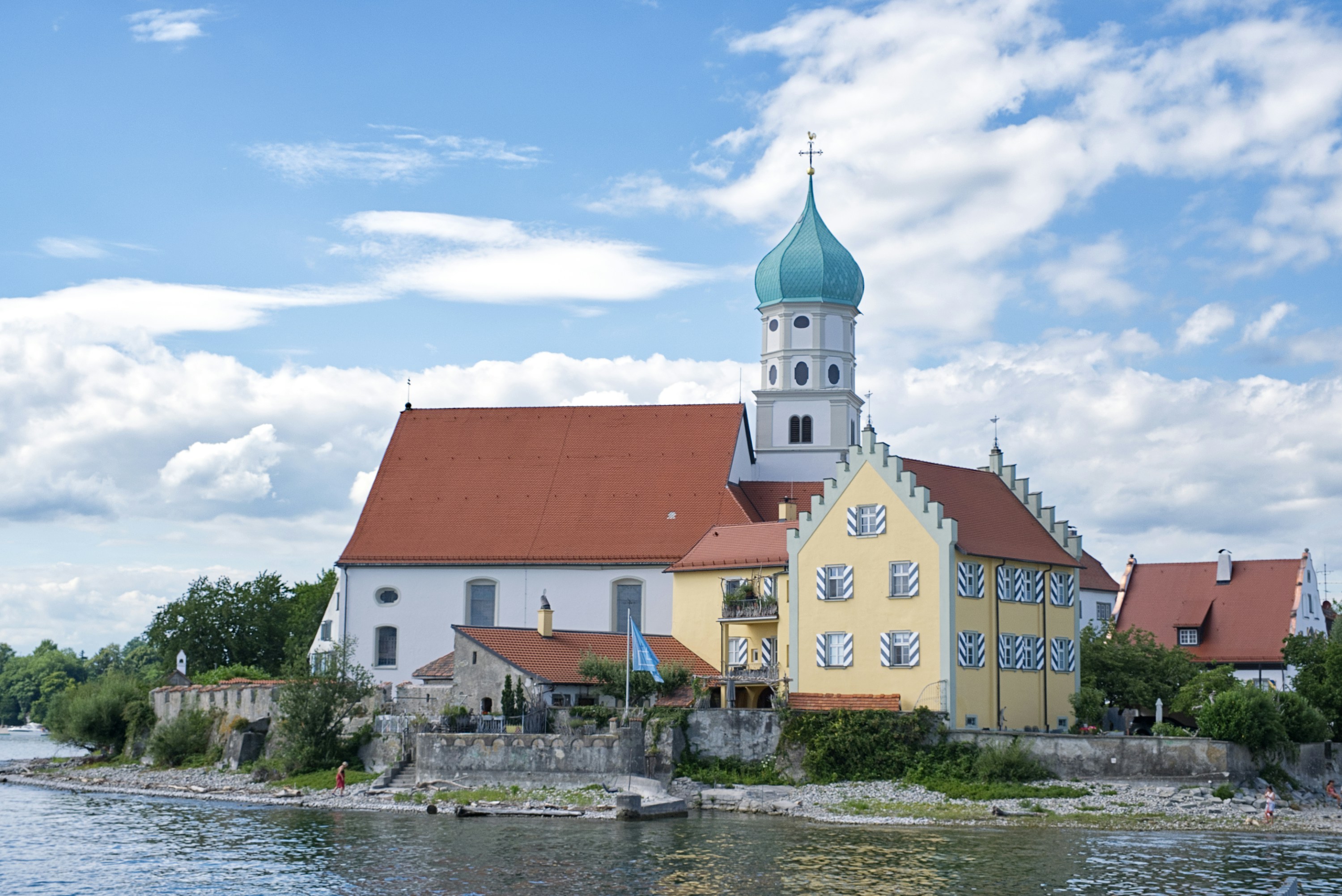 Colorful riverside buildings with a turquoise-domed church rise from the bank, reflection shimmering on the water and a bright sky above.