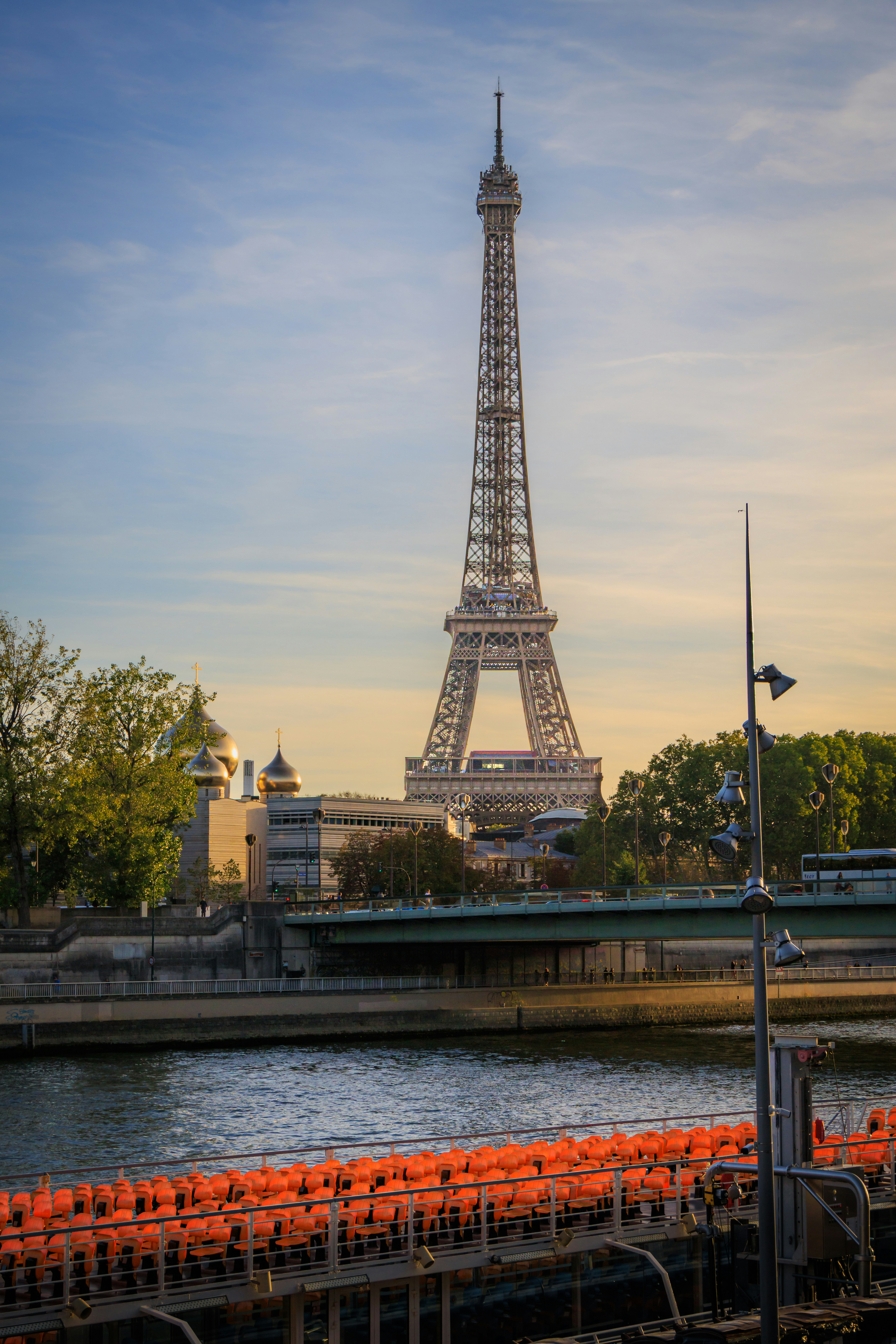 Golden-hour view of the Eiffel Tower along the Seine, with a foreground of red-orange rowers’ seats and a riverside walkway. The scene emphasizes architectural silhouette and tranquil water.
