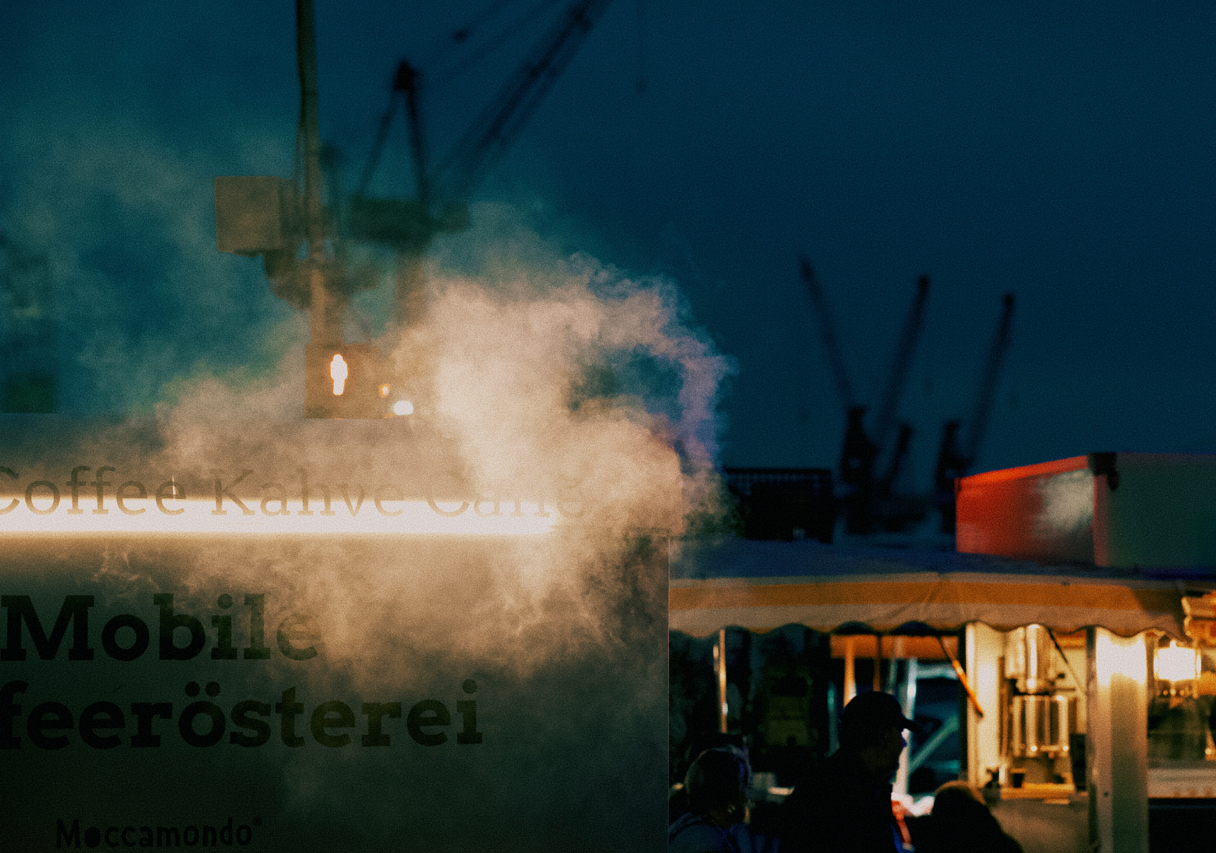 Steam over a night market sign at dusk as a subtle glow reveals silhouettes