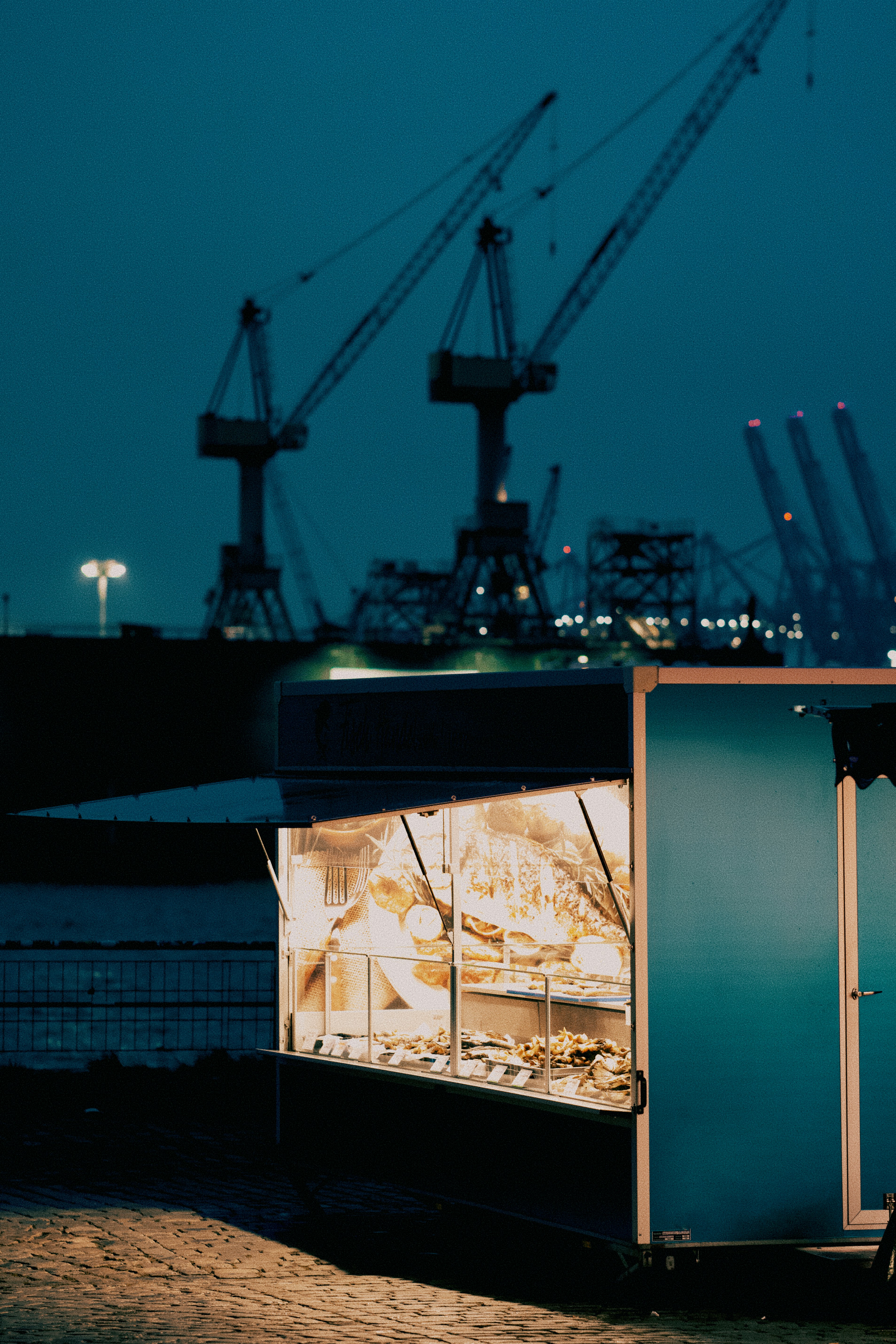 Blue-hour scene of a lit food kiosk in the foreground with an industrial port and cranes fading into the distance.