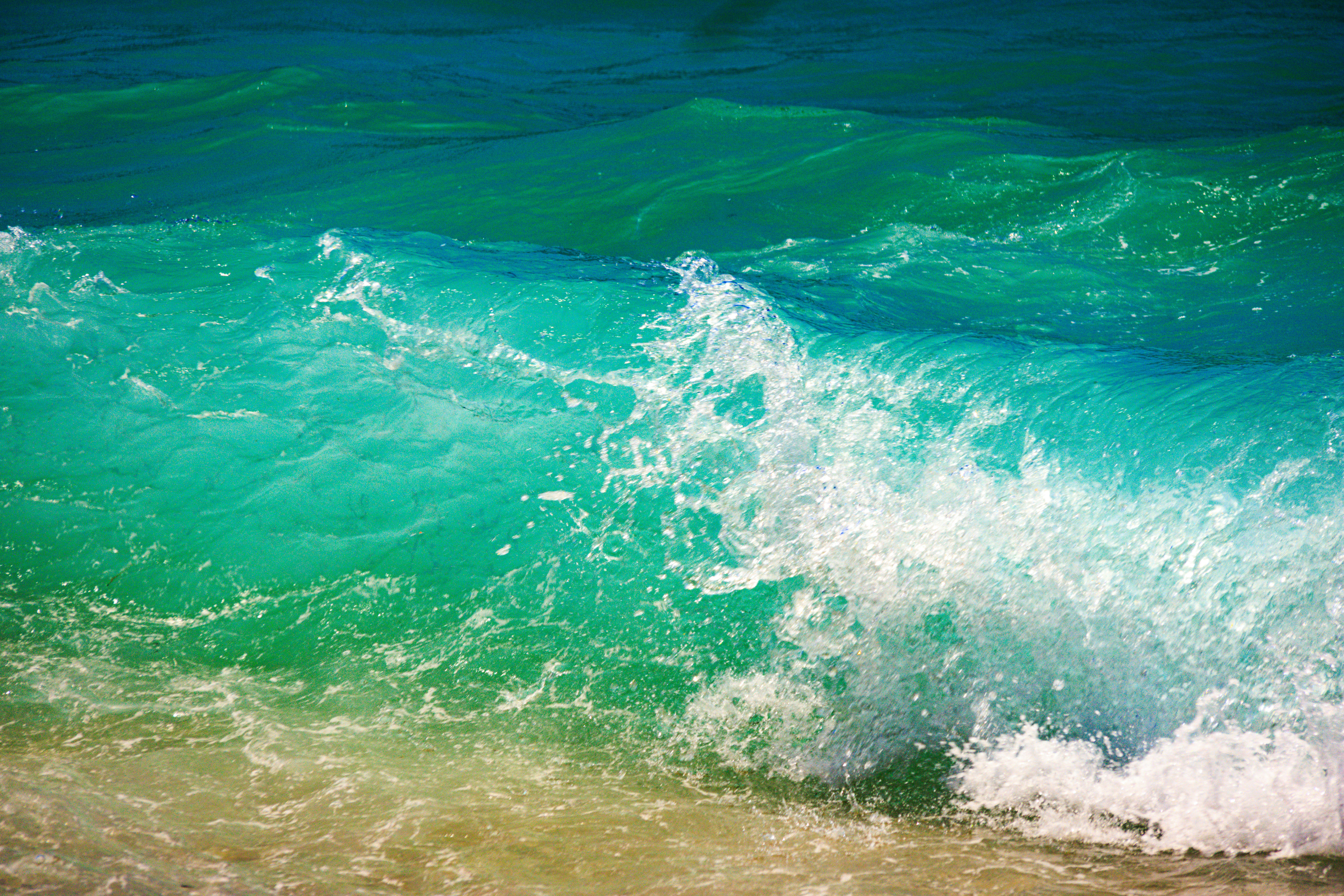 A turquoise ocean wave breaking over a sandy shore with white sea foam