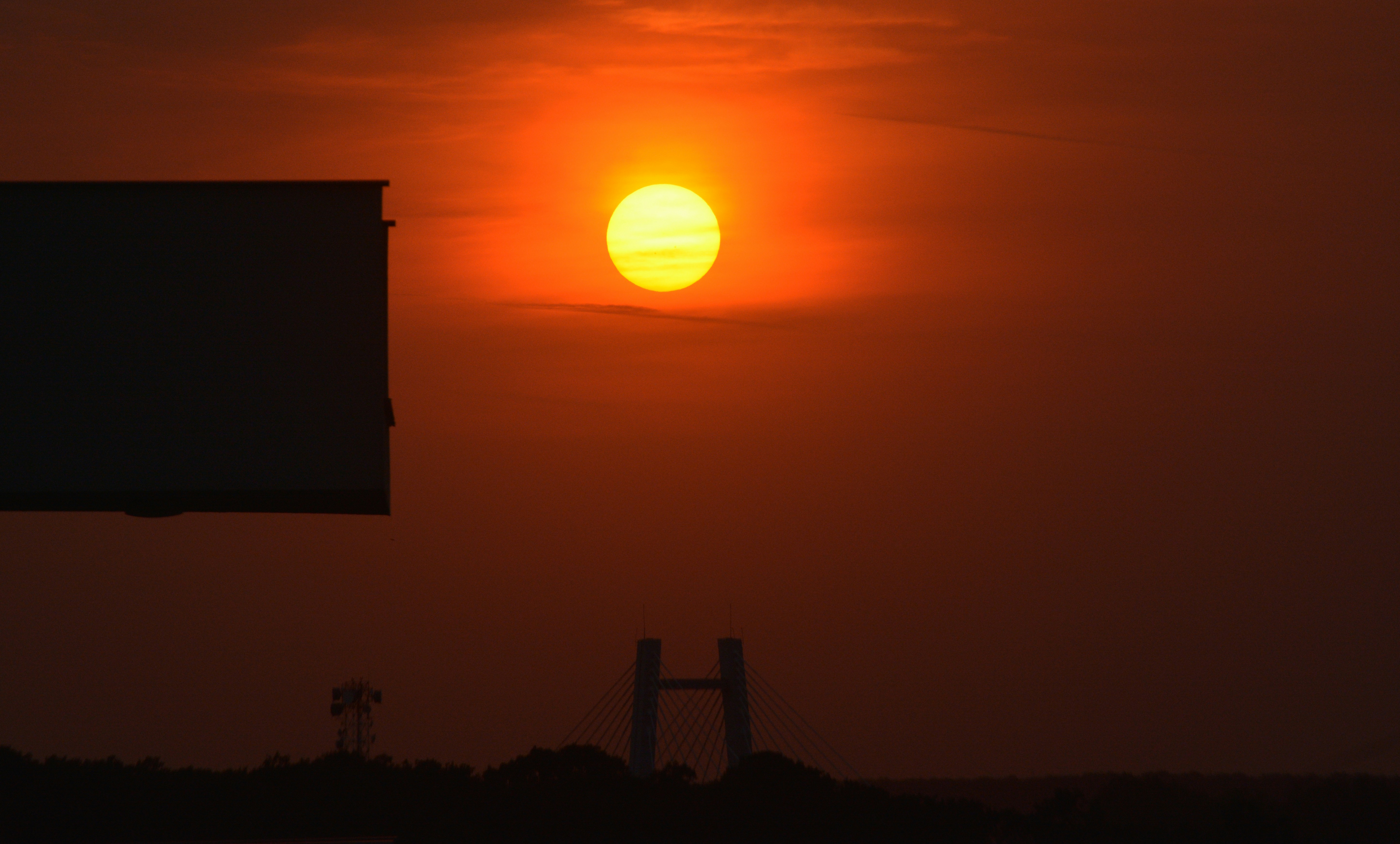 Golden sun sinks behind a dark skyline, casting warm orange hues over a silhouetted rooftop and distant bridge; a minimal urban landscape at dusk.