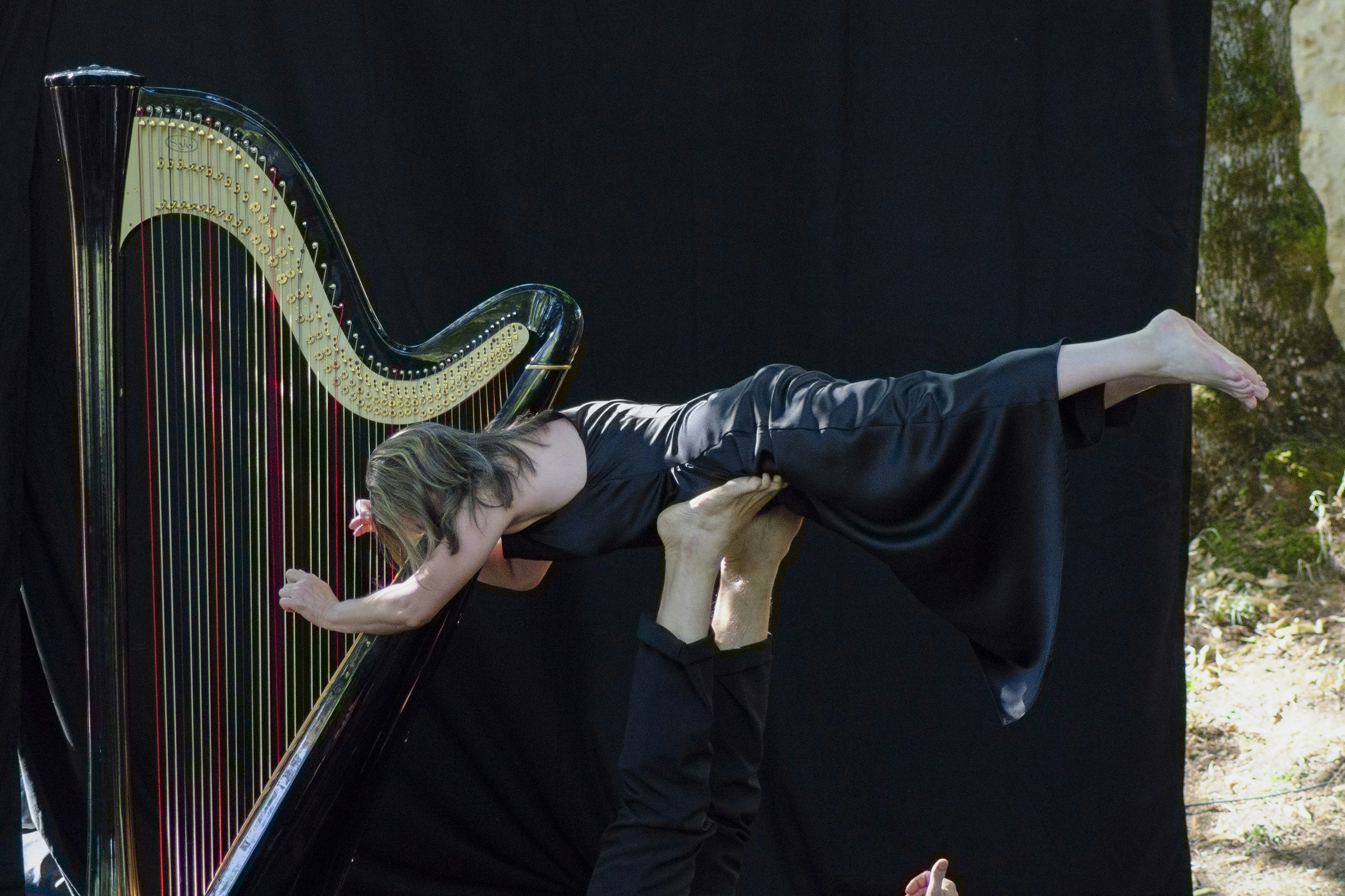 Harper leaning over a grand harp as an unseen performer lifts her leg in a dramatic pose; the scene captures motion, balance, and a moment of performance art against a dark backdrop.
