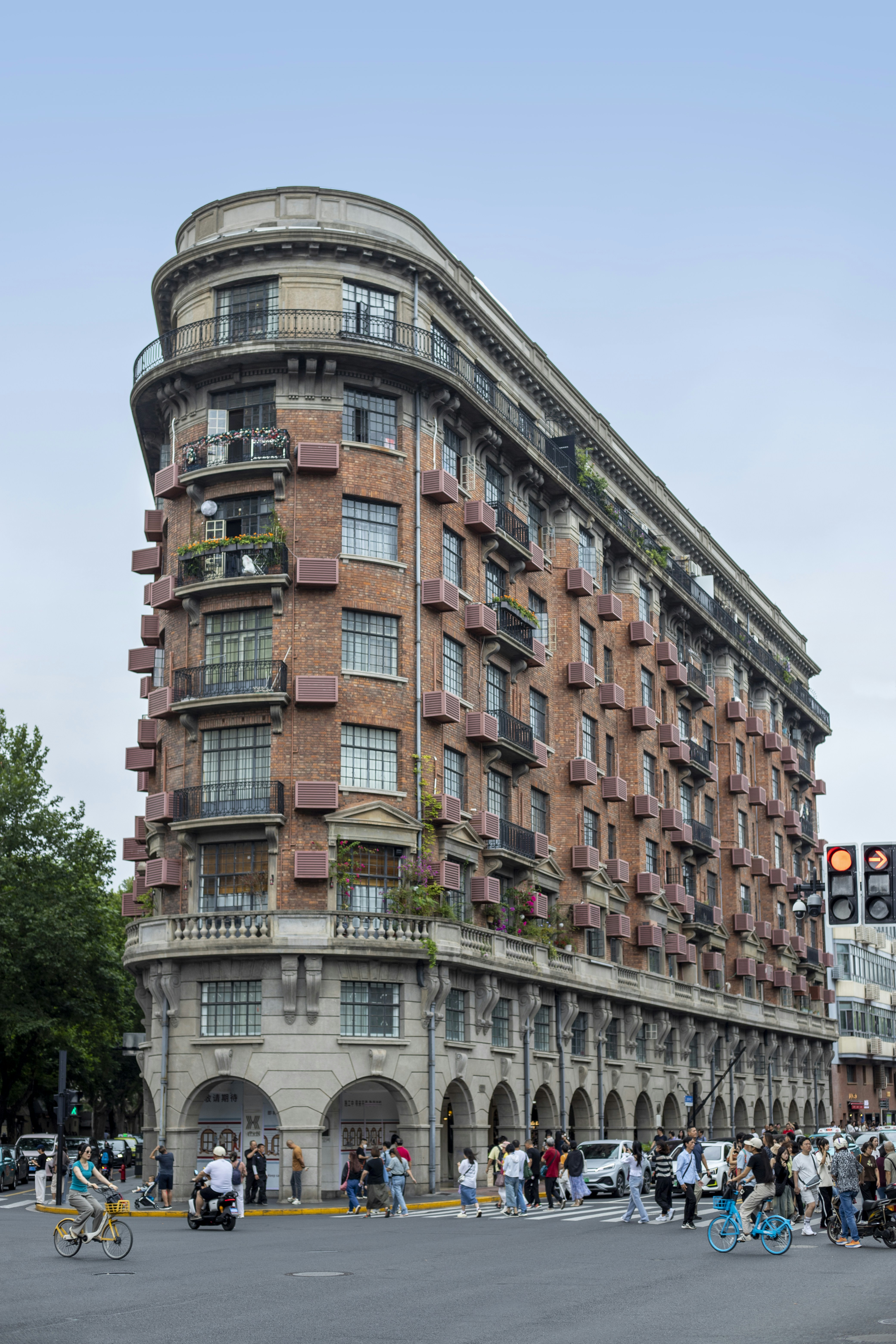 Brick residential building with rounded corner and balconies dominates a crowded urban intersection; pedestrians and cyclists fill the street below.