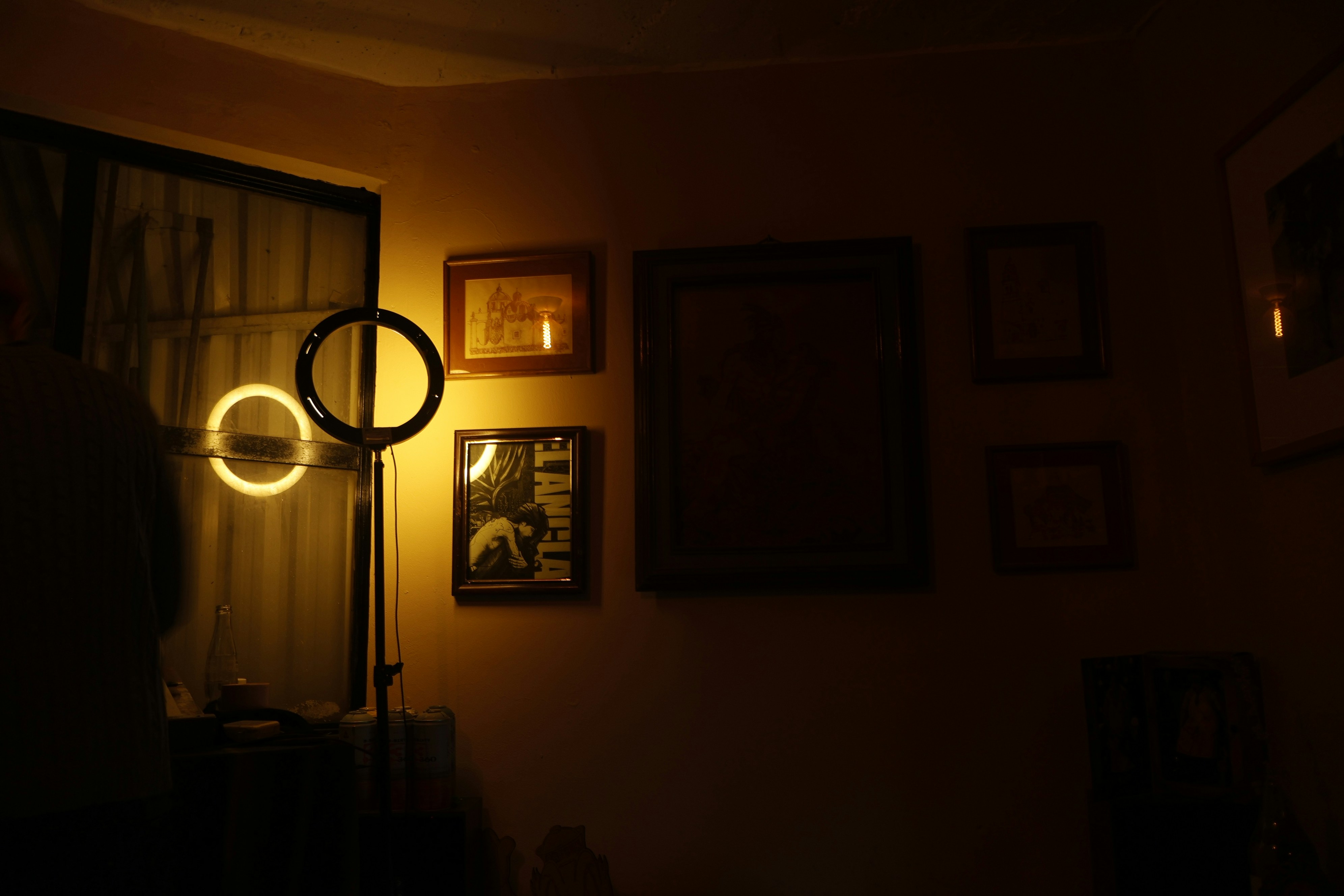Low-light interior scene featuring a ring light on a stand casting warm glow onto framed artworks. The setup suggests a small studio or filming space with soft, ambient illumination and shadows across a cluttered wall.