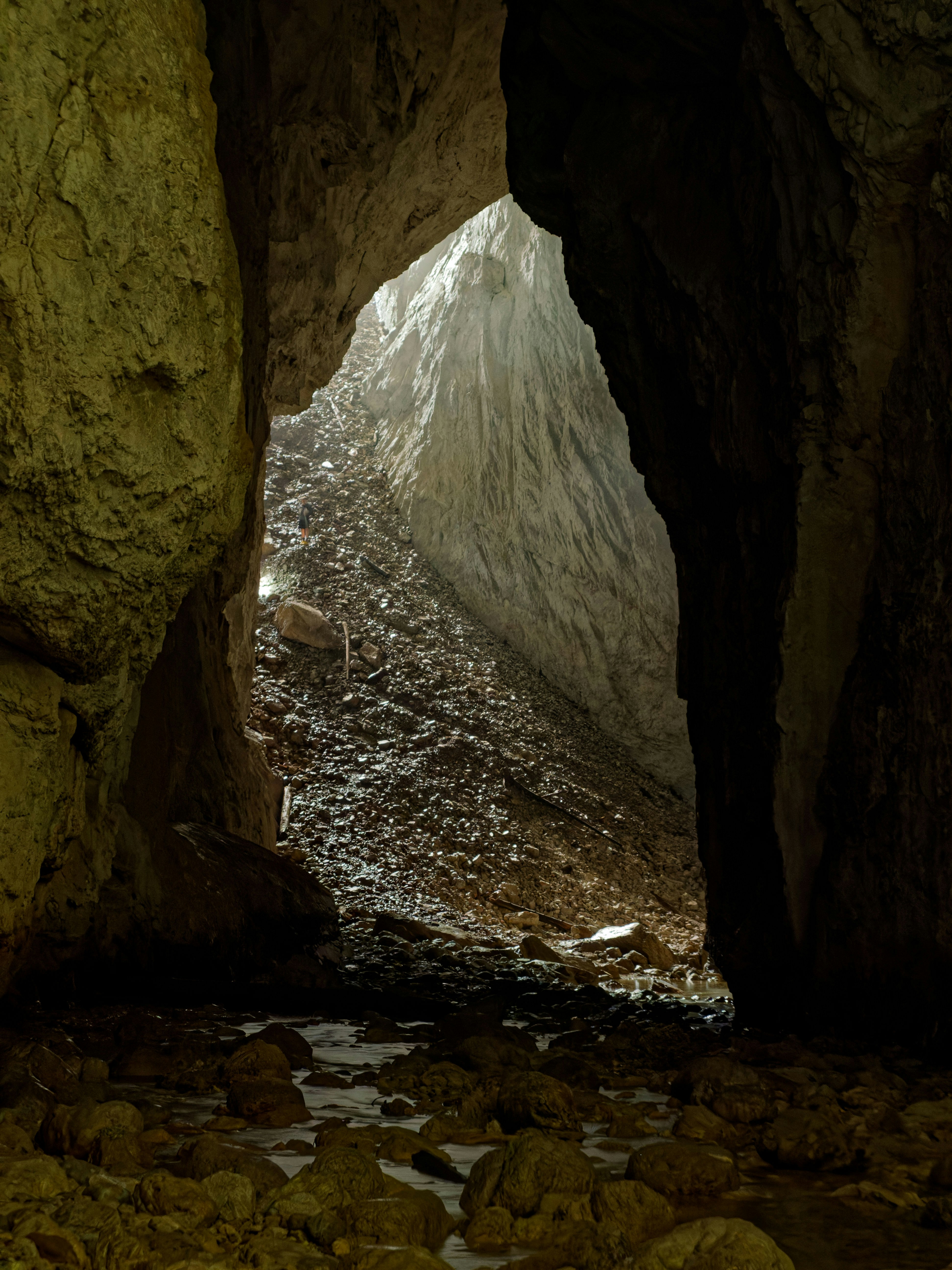 A dim cave opening leads to a sunlit passage, with rough rocky walls framing a narrow, rocky floor and a misty glow at the far end.