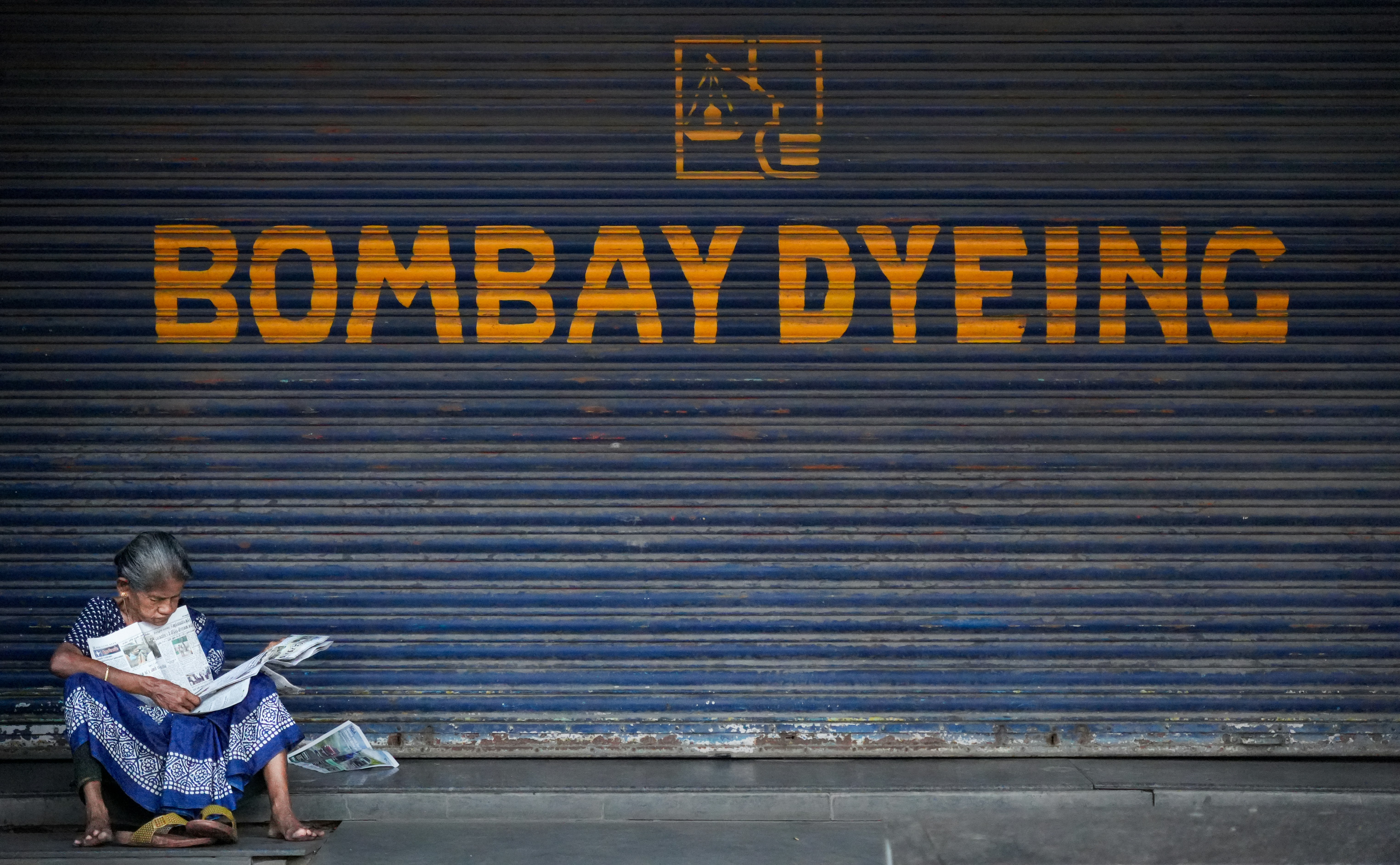A lone elderly person sits on a curb reading on a blue storefront shutter, outerwear folded, suggesting a quiet moment in an urban setting. A sign reading “BOMBAY DYEING” looms above, establishing location and mood.