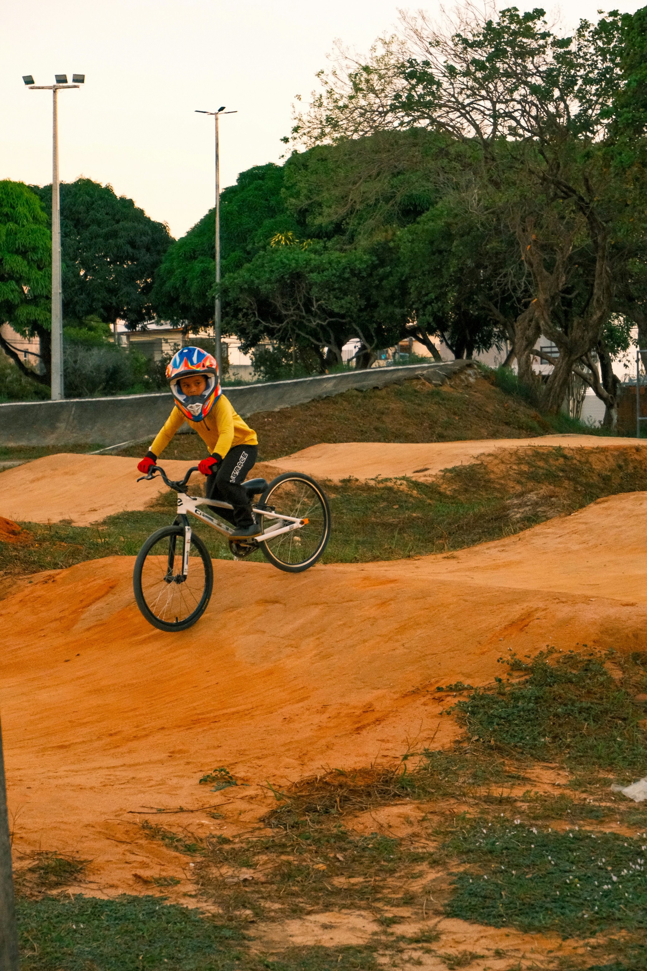 Youth rider in a yellow jersey balances a BMX across orange dirt mounds at a skate park; trees and light poles provide a suburban backdrop.