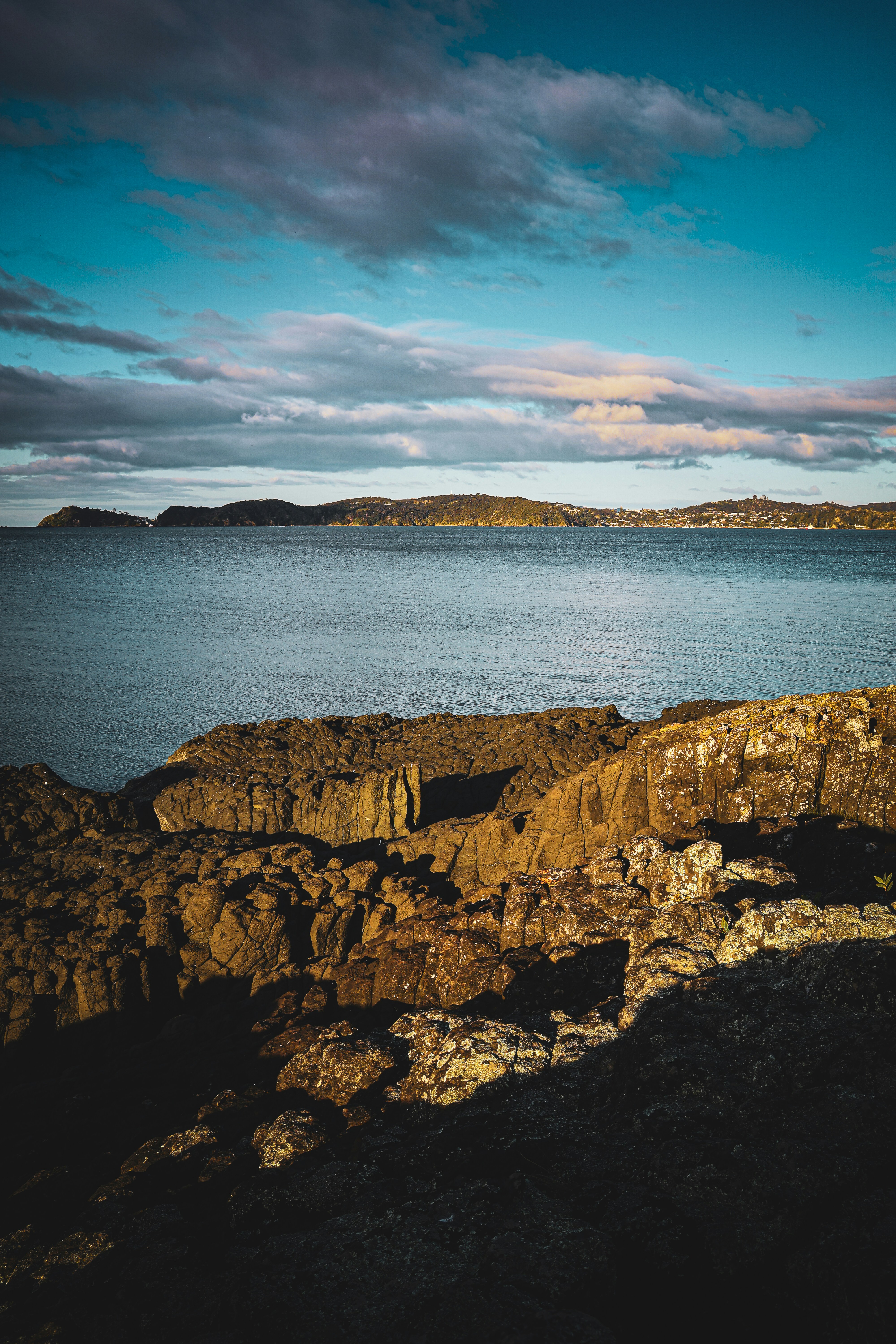 Rocky shoreline bathed in warm light meets calm sea under a bright sky; the scene captures rugged terrain leading to distant isles. This is a landscape photograph.