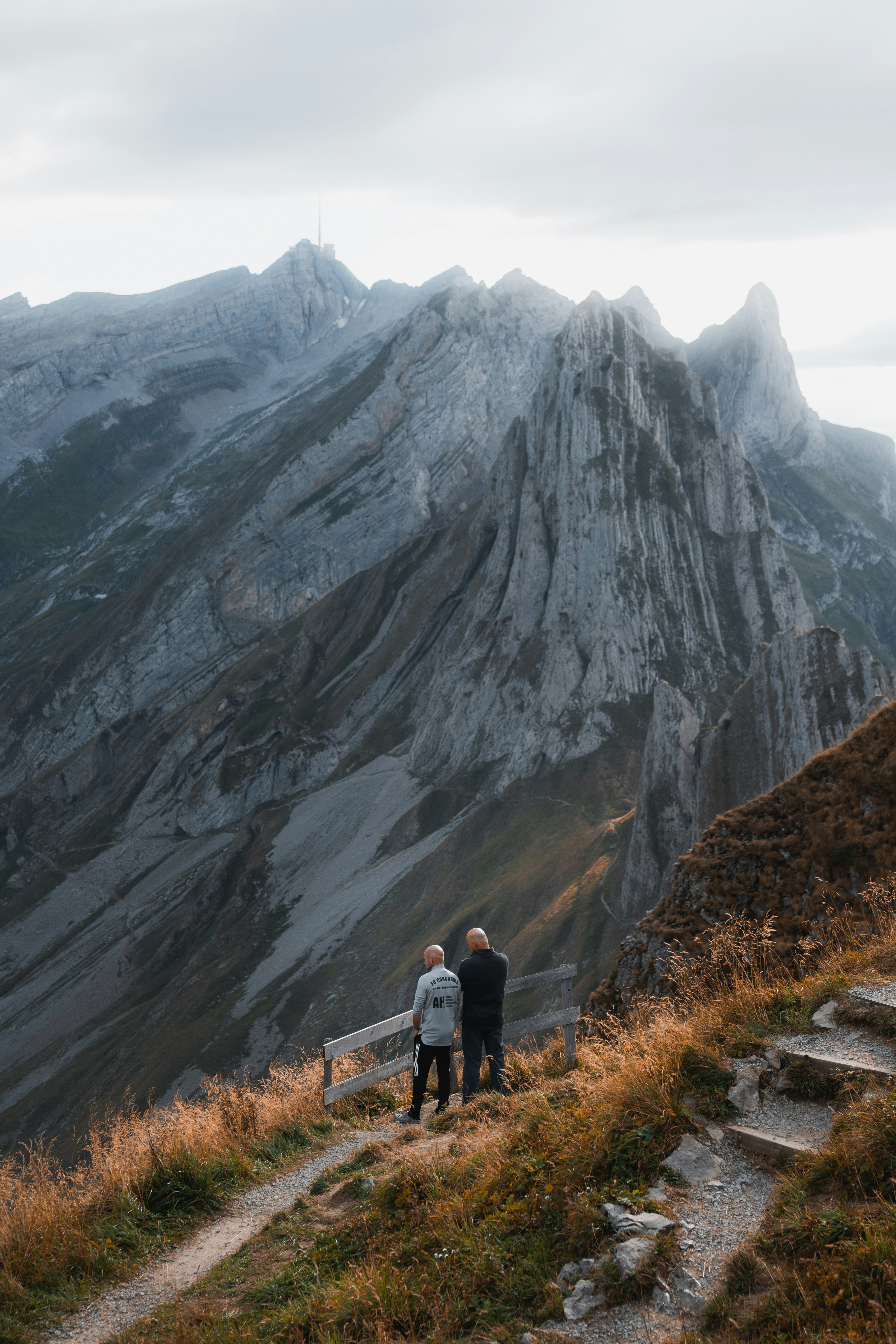 Two hikers at a precipice overlook beneath jagged alpine peaks