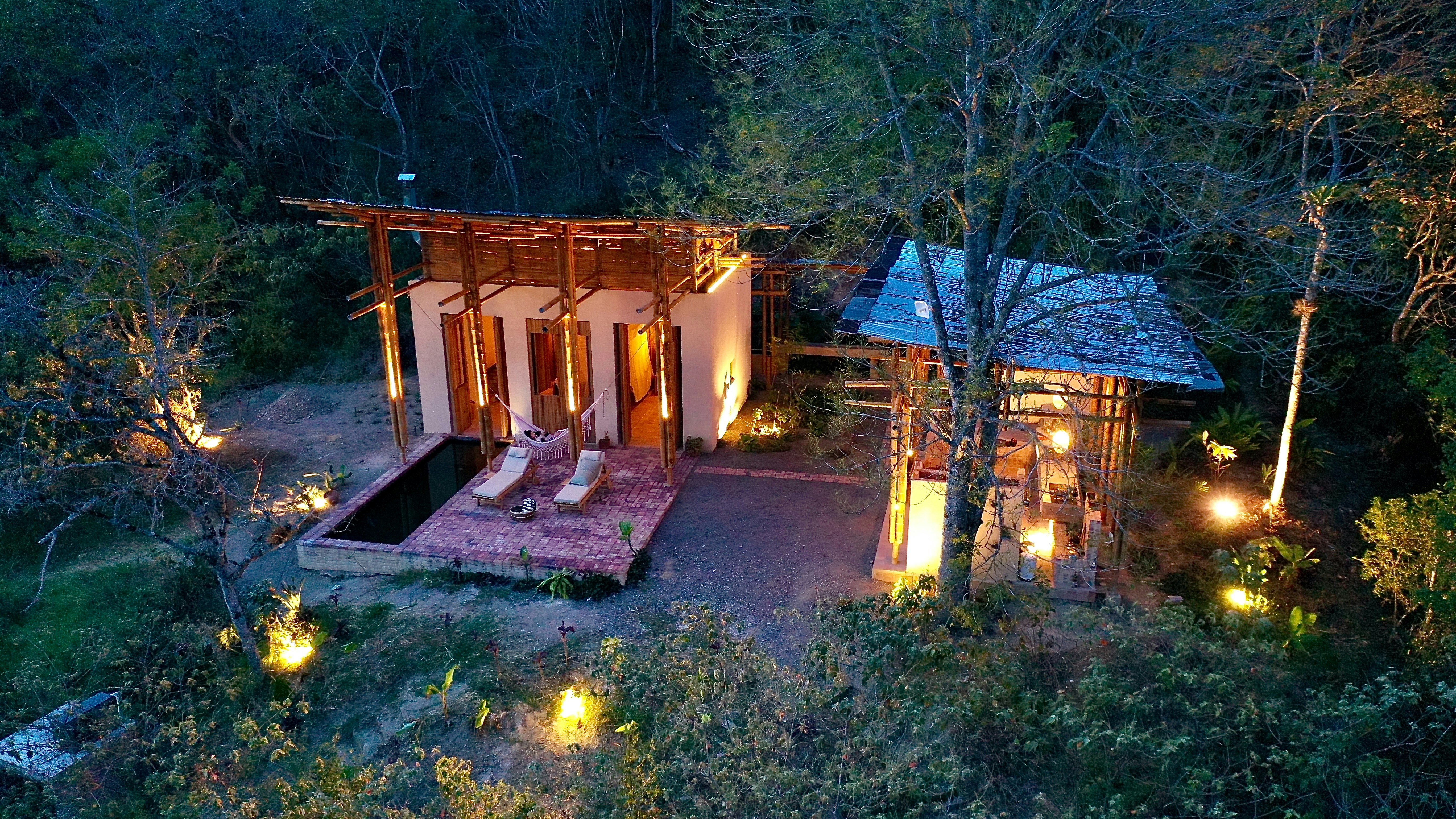 An illuminated modern retreat nestled among trees at dusk, with a wooden pergola, patio, and secondary shelter glowing softly in a garden setting.