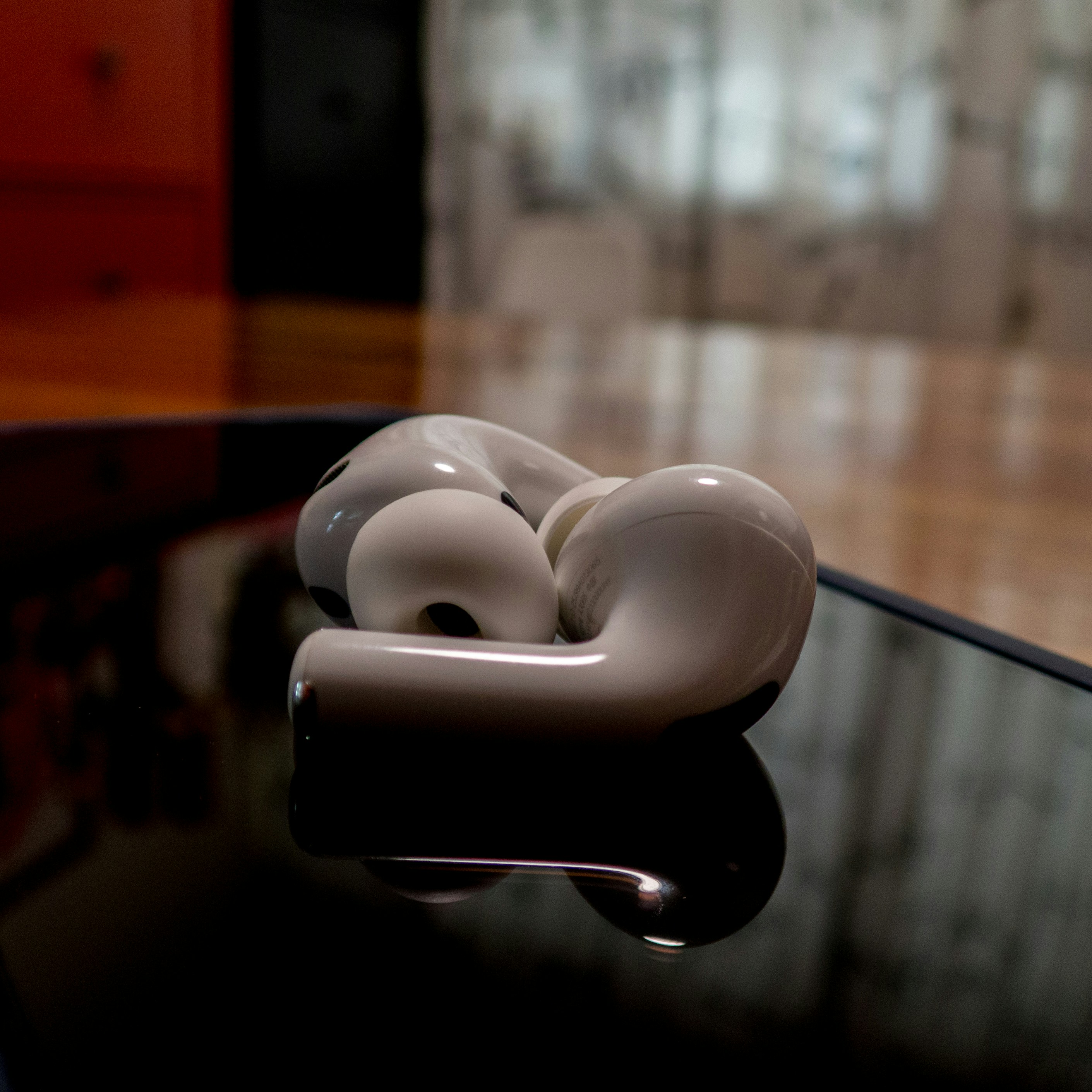 Sleek white wireless earbuds sit on a glossy dark surface, mirrored in the reflections, with a blurred indoor background. A minimal still-life focusing on shape and curve.