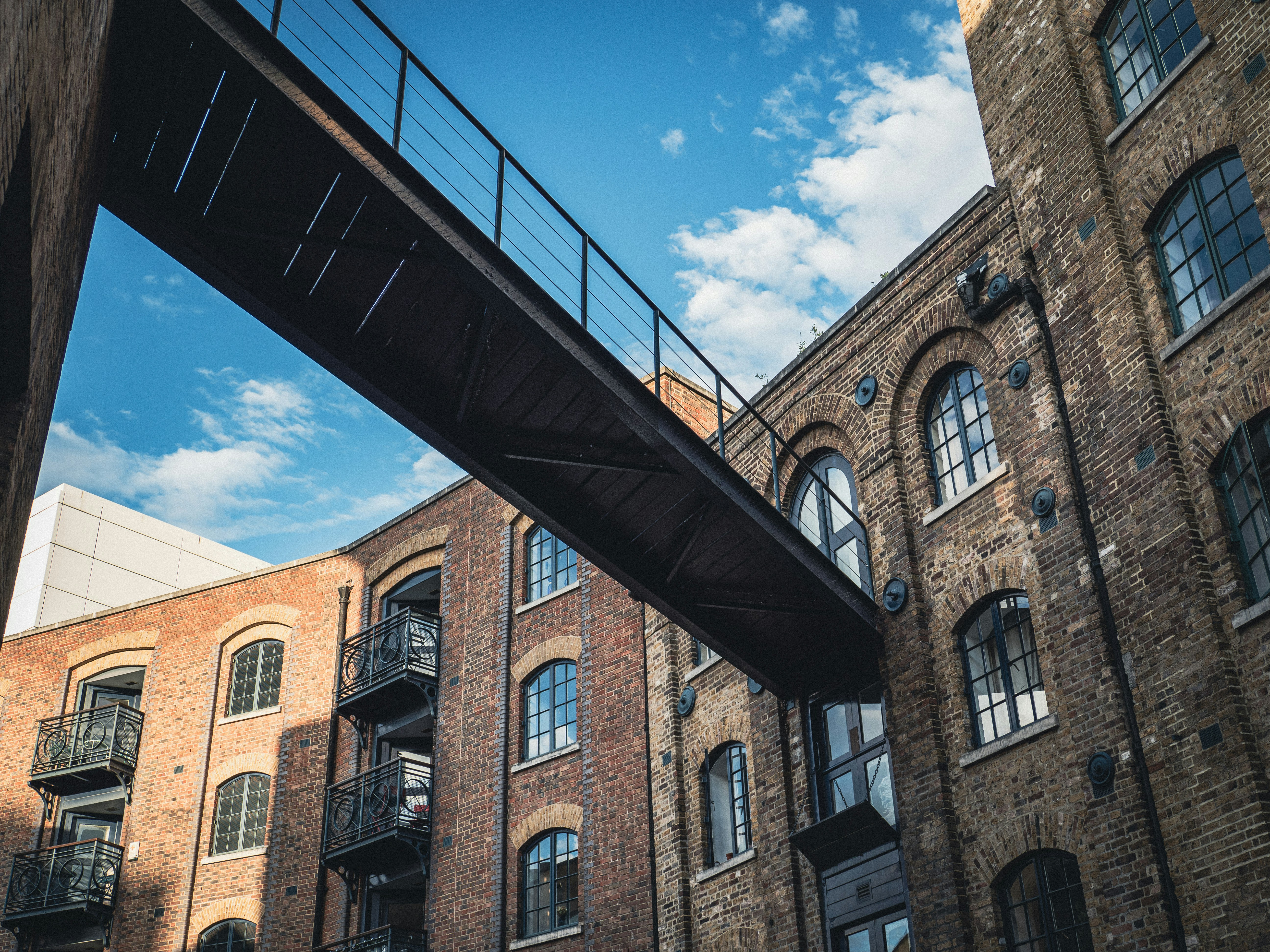 Brick warehouses with a metal skybridge linking two buildings under a blue sky; an architectural detail emphasizing urban texture and lines.