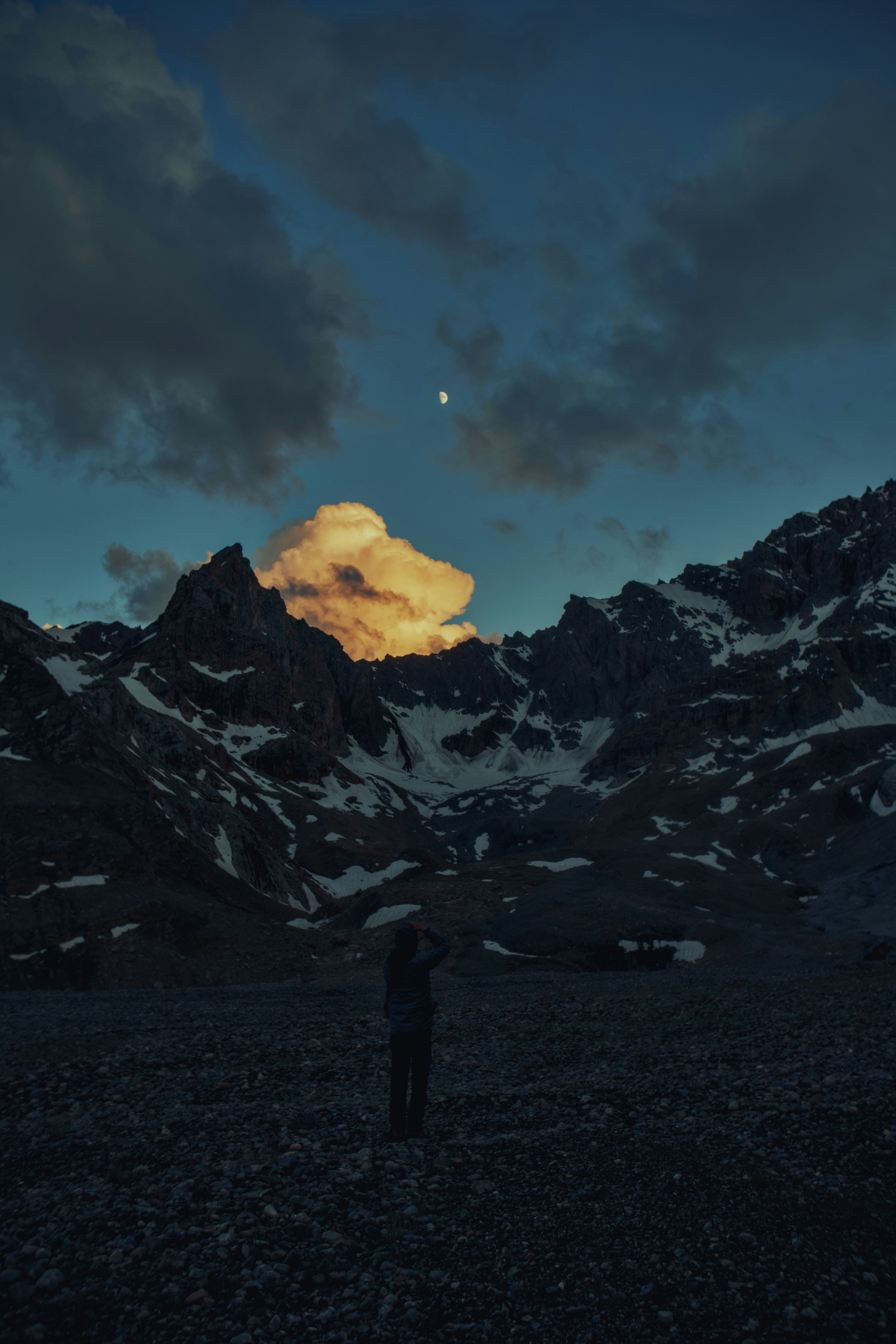 Moonlit summit: a lone figure beneath a glowing cloud over jagged peaks