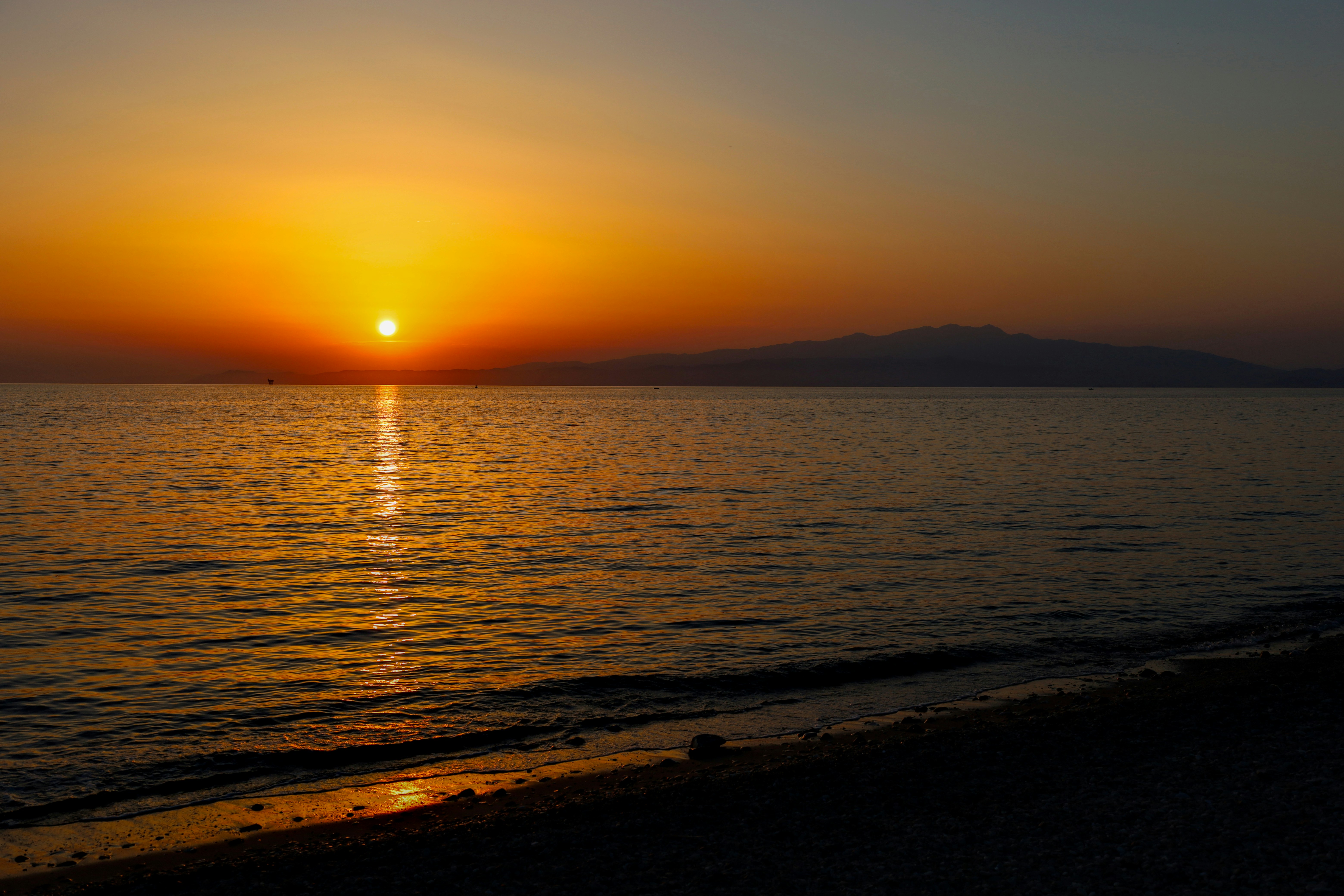 Warm sunset over calm sea with the sun low on the horizon and a dark shoreline in the foreground. A tranquil coastal scene emphasizing golden reflections on gentle waves.