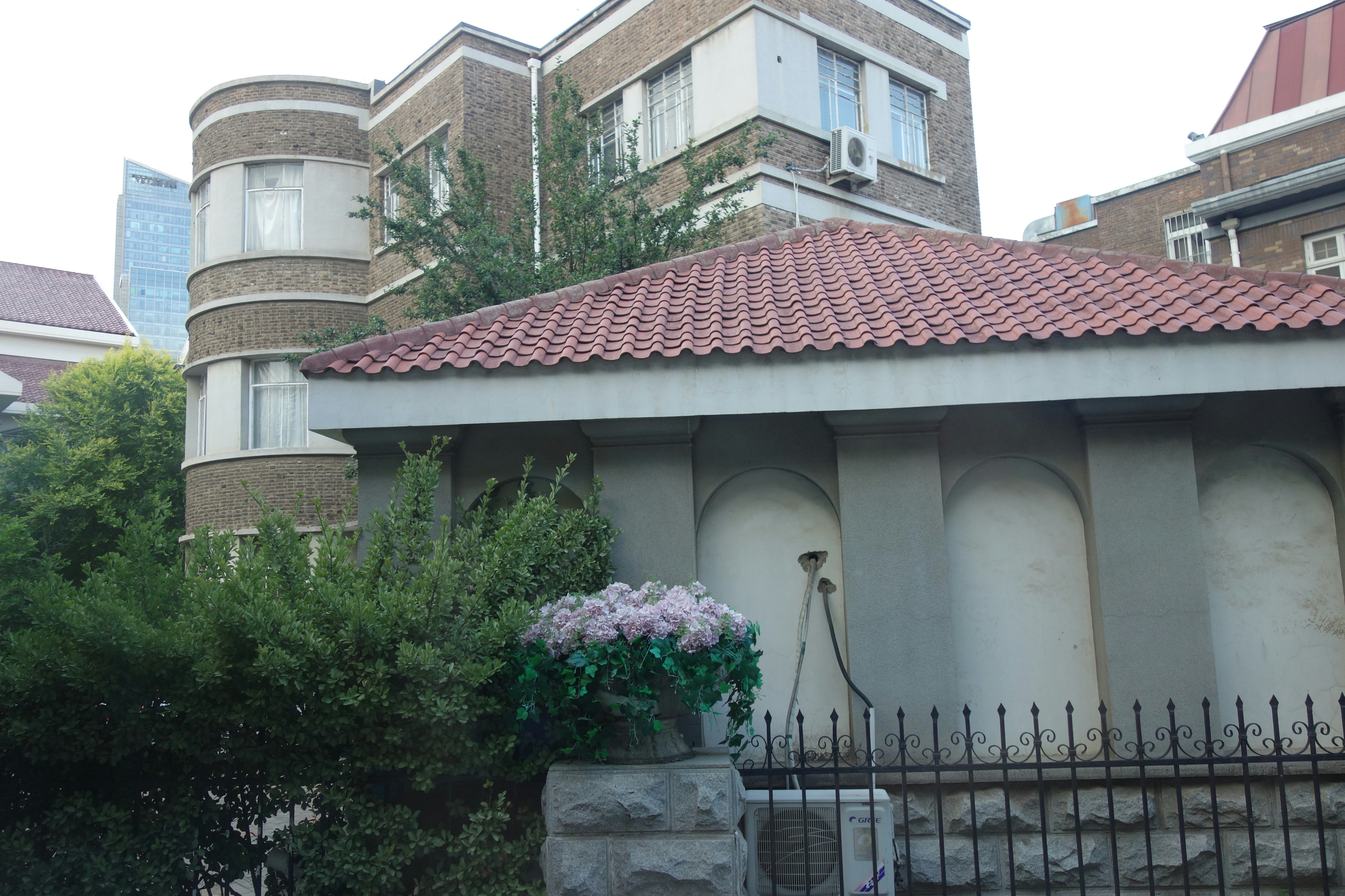 Corner view of a small architectural complex with a red-tiled roof, stone wall, and blooming shrubbery in the foreground, presenting a quiet courtyard scene.