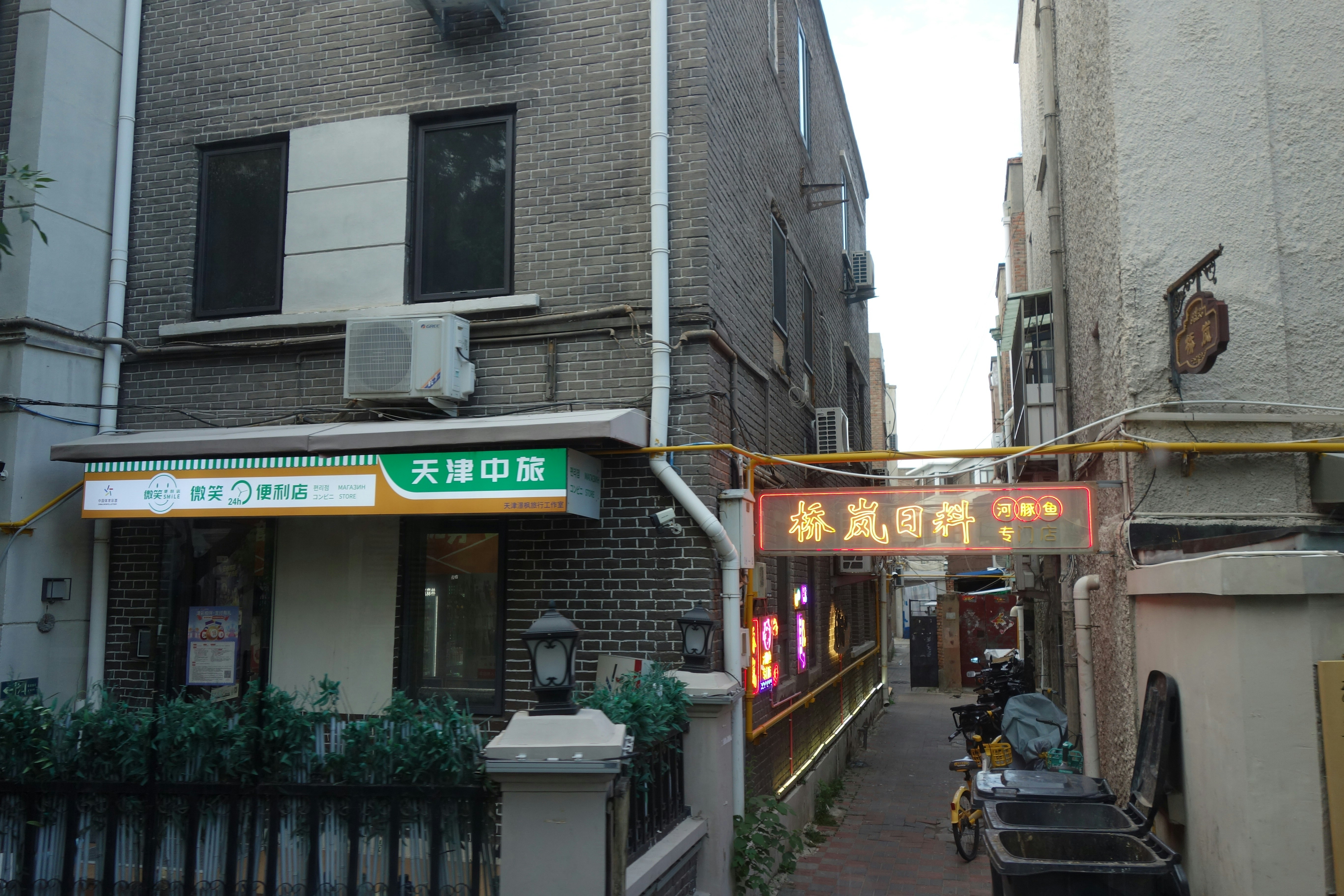 Narrow urban alley with storefronts, hanging signs, and a scooter at the end; a pedestrian-lit passage framed by brick buildings and warm lantern glow.
