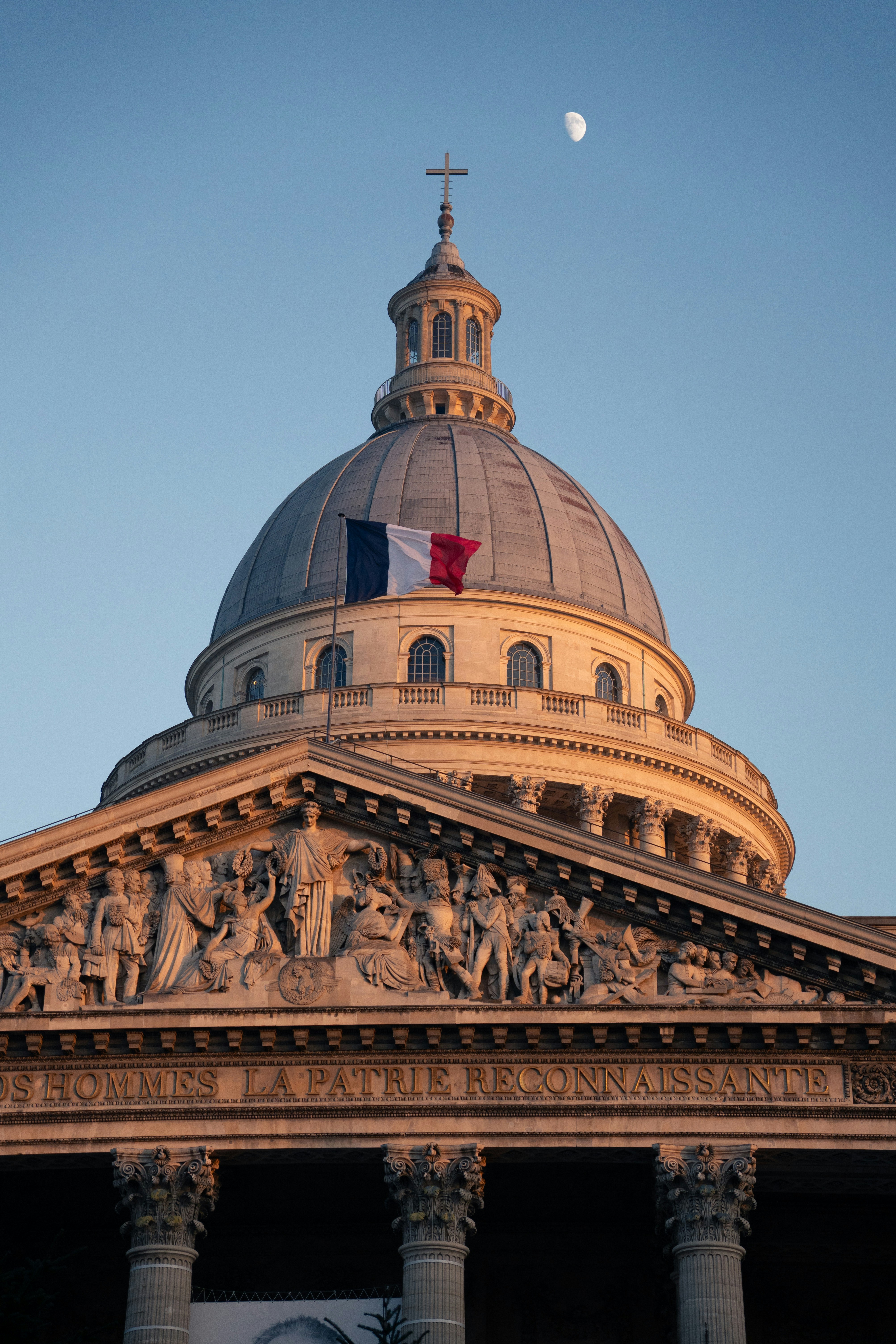Dome of a grand cathedral with a flag at the base and sculpted friezes below, set against a pale blue sky with a small moon visible above.