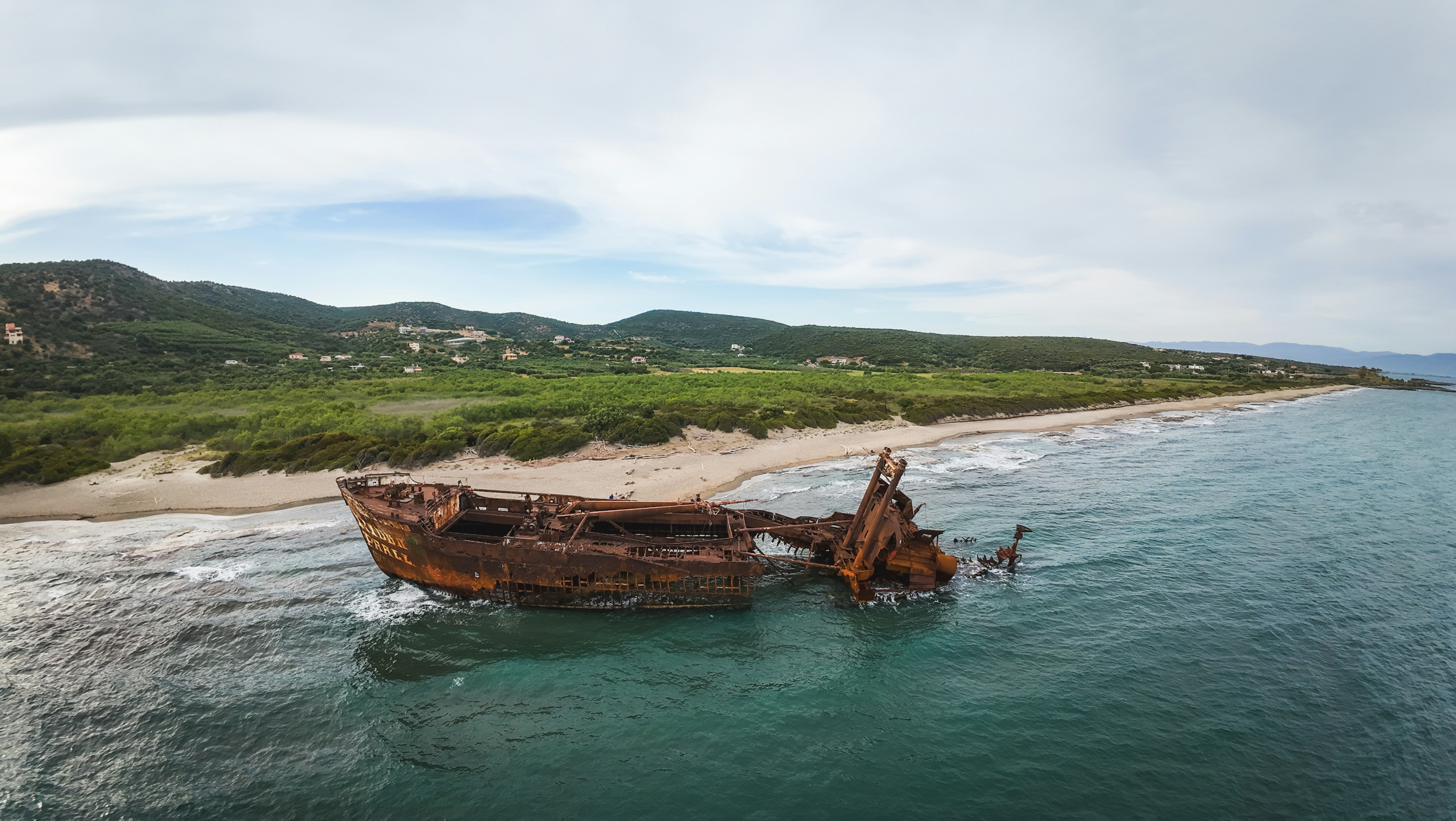 Rusted shipwreck stranded on a sandy, green shoreline with calm turquoise water and distant hills. Aerial view highlights the vessel's decay and the surrounding coastal landscape.