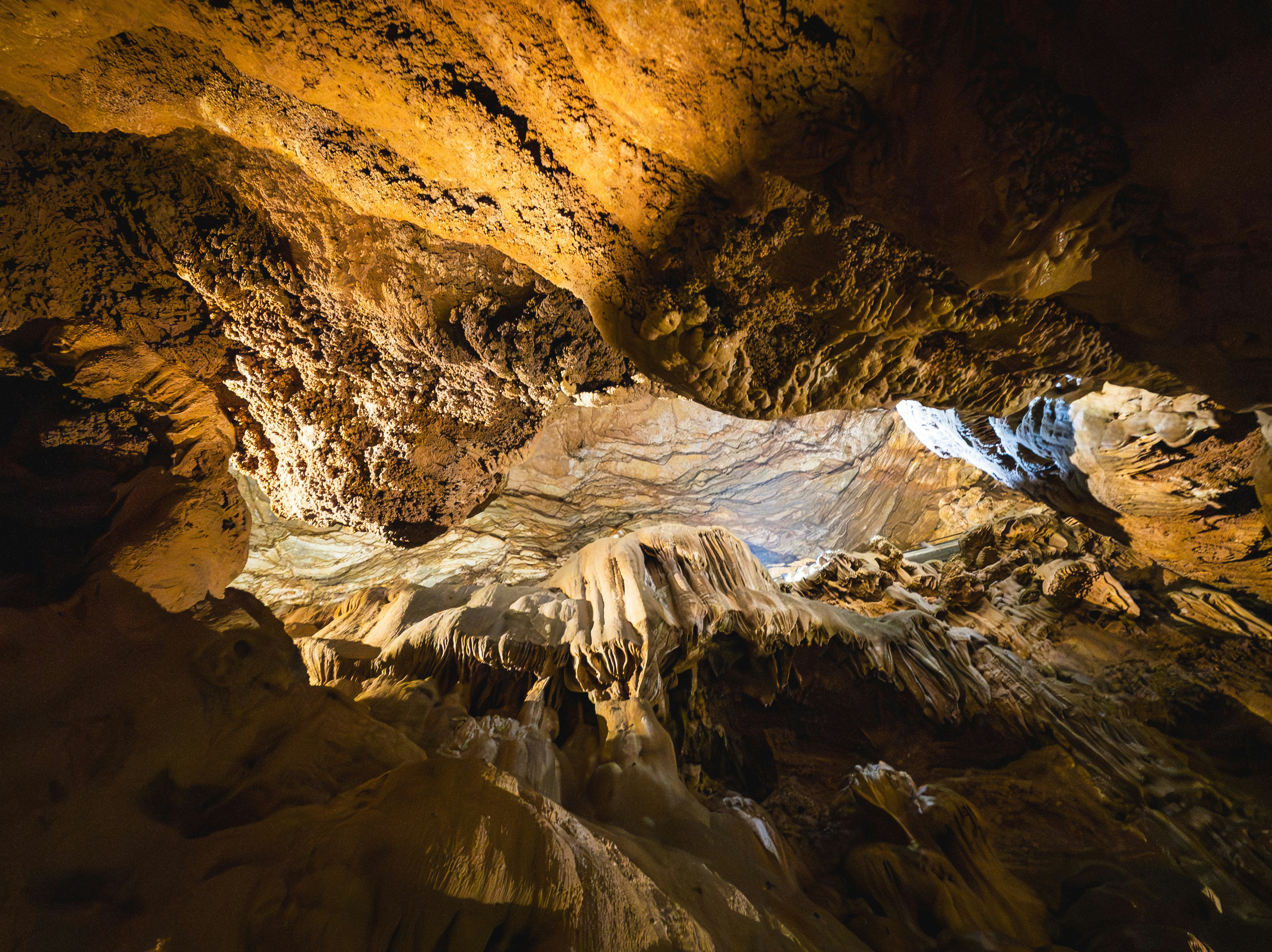 Dimly lit cave interior with stalactites and stalagmites bathed in warm and cool light, creating textured rock formations and a distant opening. A rugged, natural abstract landscape within a subterranean chamber.