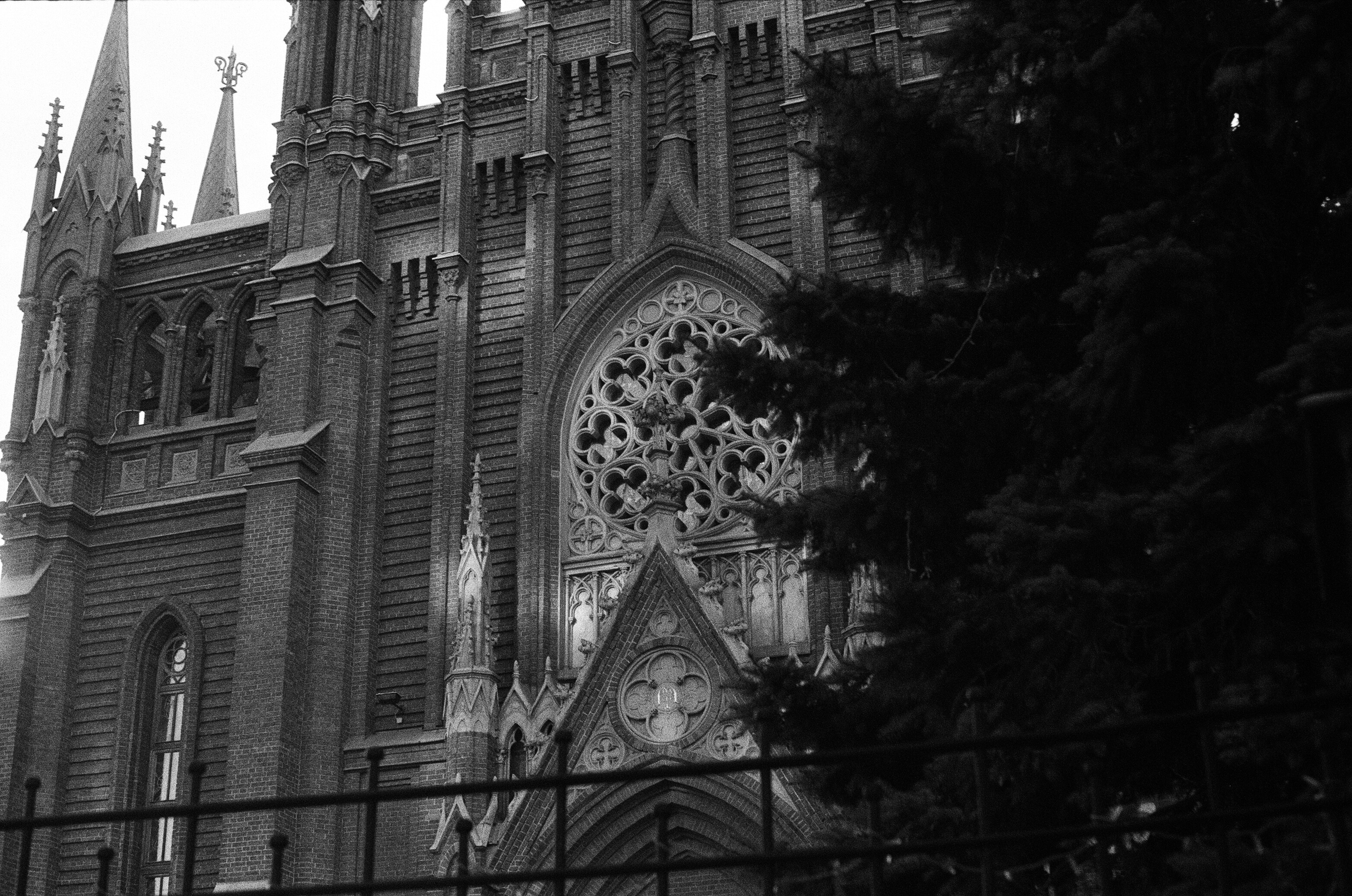 Dark, detailed church facade with a prominent rose window and spires, viewed behind a metal fence; high-contrast black-and-white rendering emphasizes stone texture and architectural motifs.
