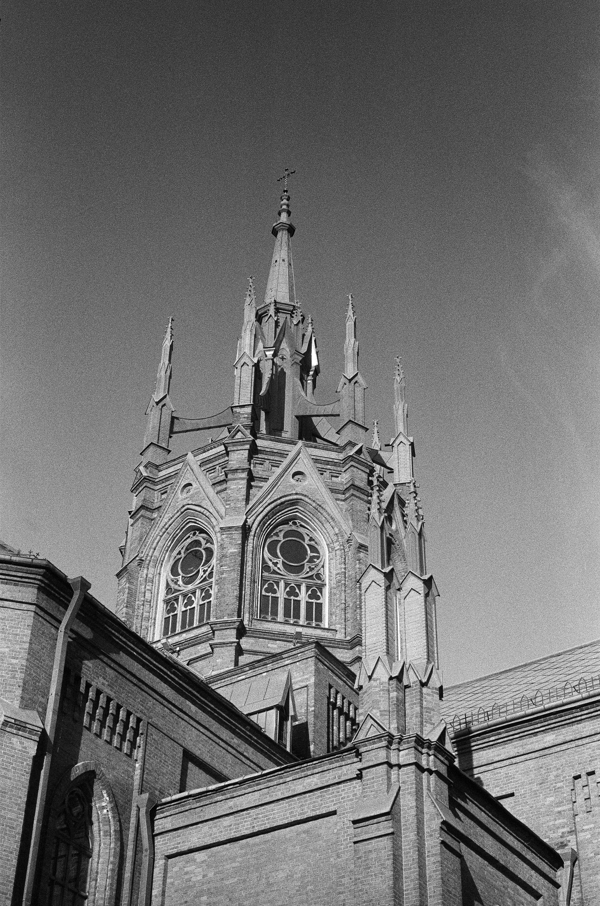 A black-and-white view of a historic cathedral-like brick tower with pointed arches and intricate window tracery, rising above foreground structures.