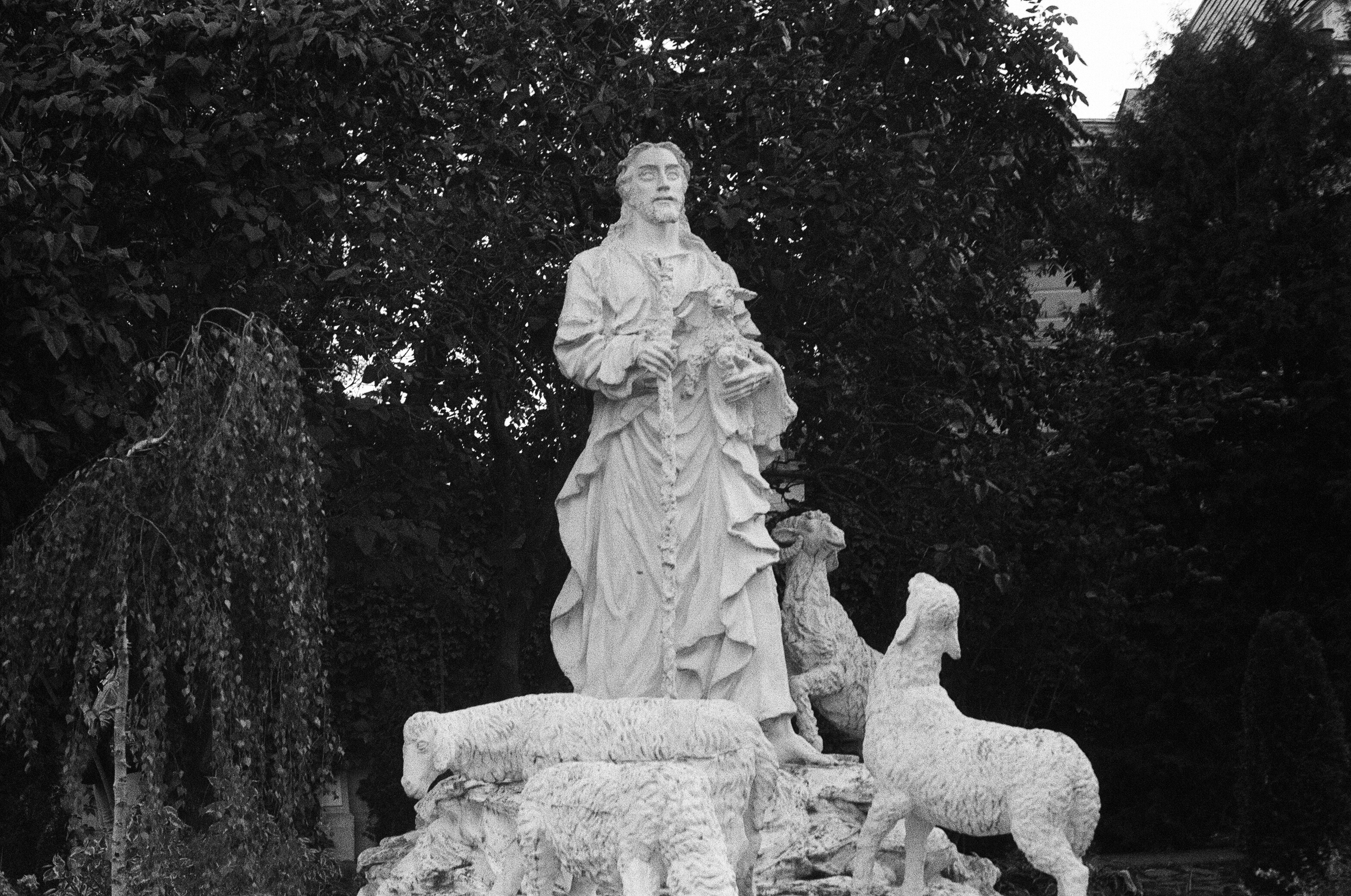 Stone statue group atop a rocky pedestal with grazing animals in a garden setting, captured in monochrome to emphasize texture and form.