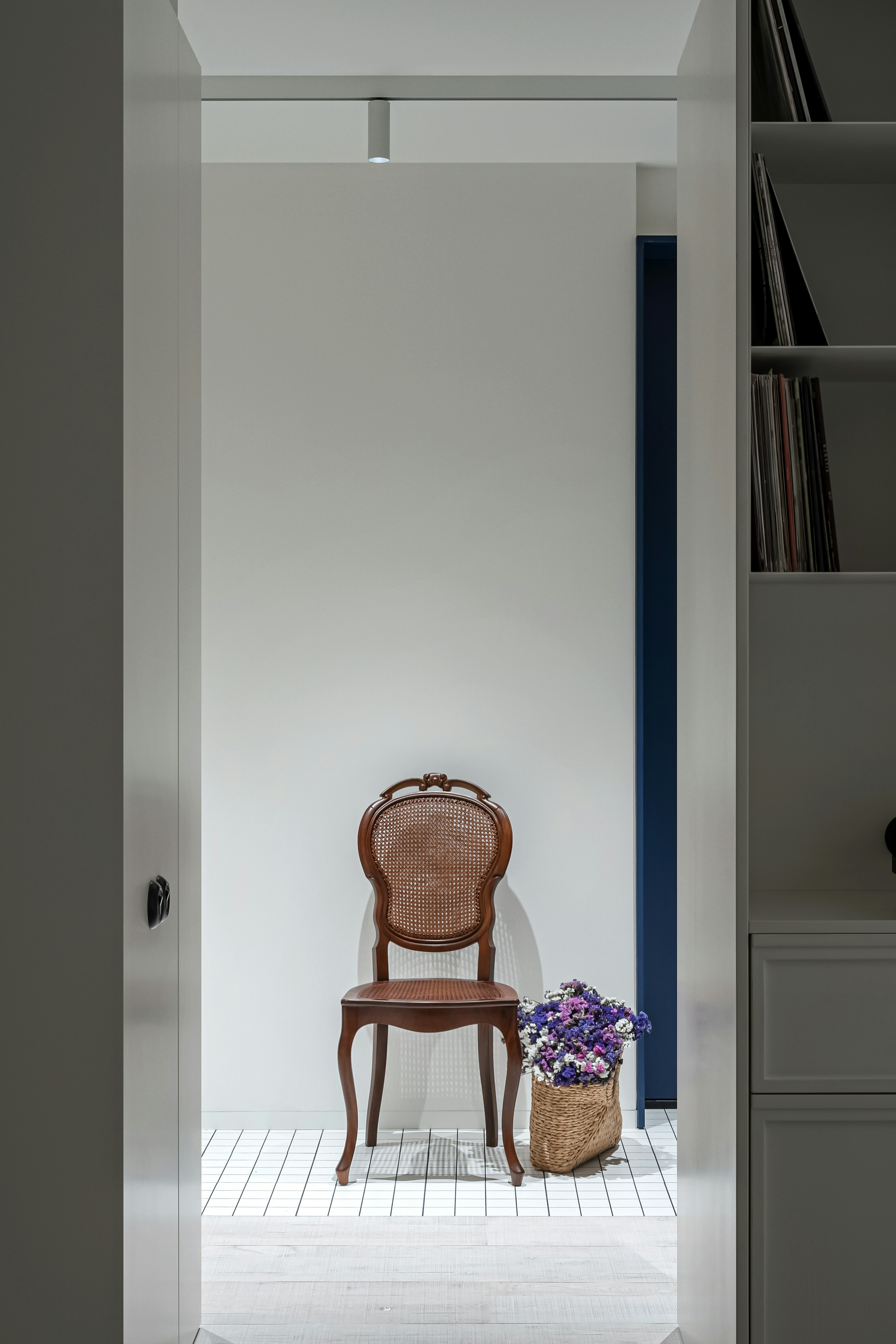 A vintage cane-back chair and a woven basket of purple flowers create a quiet focal point in the minimalist hallway of a Kyiv apartment. White cabinetry, tiled flooring and a deep blue doorway frame the scene. Interior design by PAINTIT; photography by Andriy Shurpenkov.
