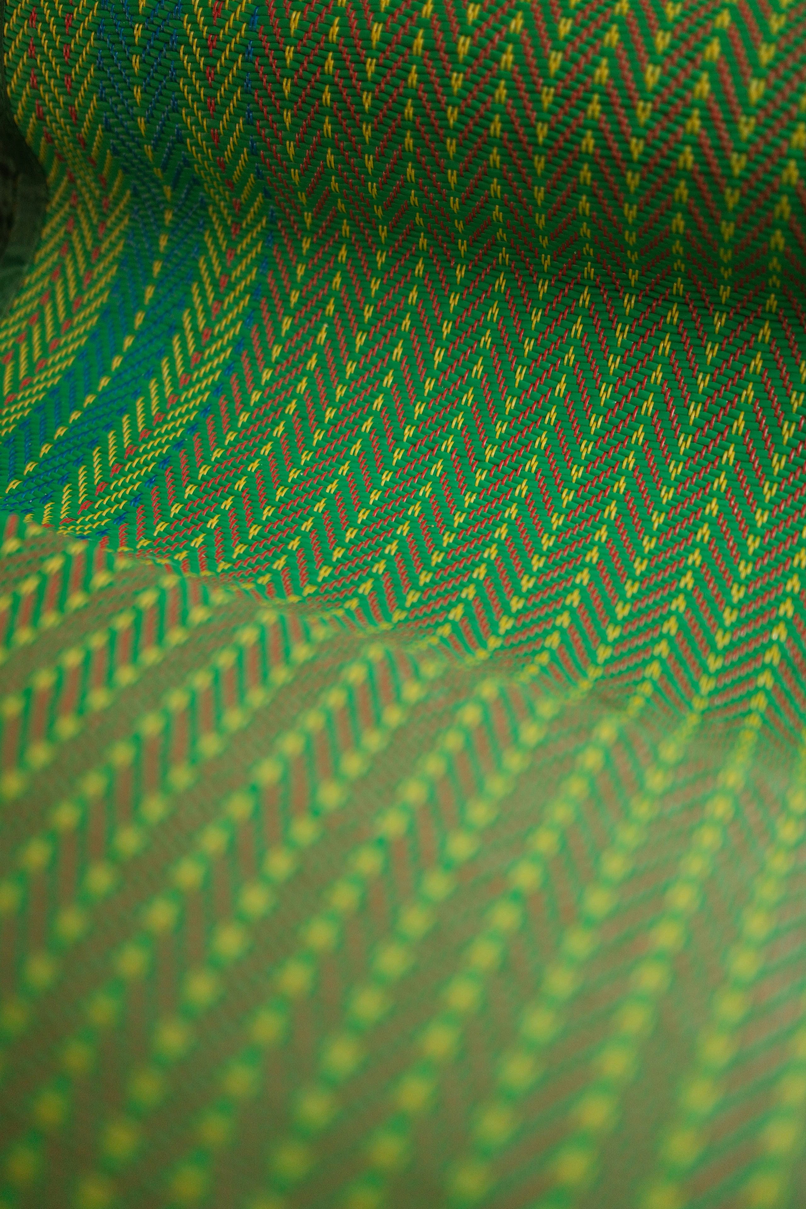 Close-up of a curved, textured green-and-gold woven fabric pattern with repeating chevron zigzags. The image highlights the detailed fabric weave and color texture without a human subject.
