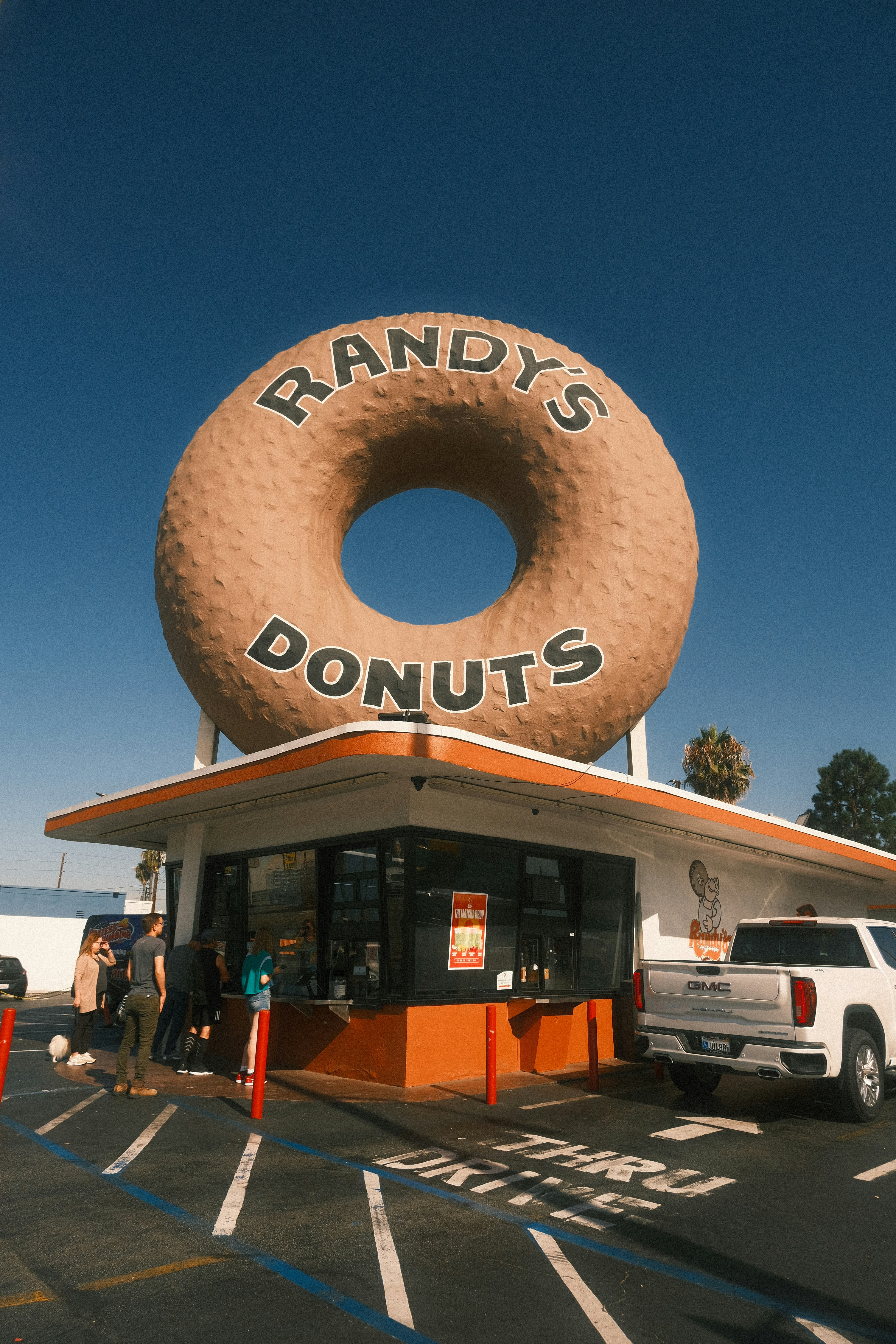Bright blue sky frames the oversized donut sign atop Randy's Donuts, with customers and a white pickup in the foreground at the colorful stand. The scene captures a casual, street-side bakery counter and parking lot.