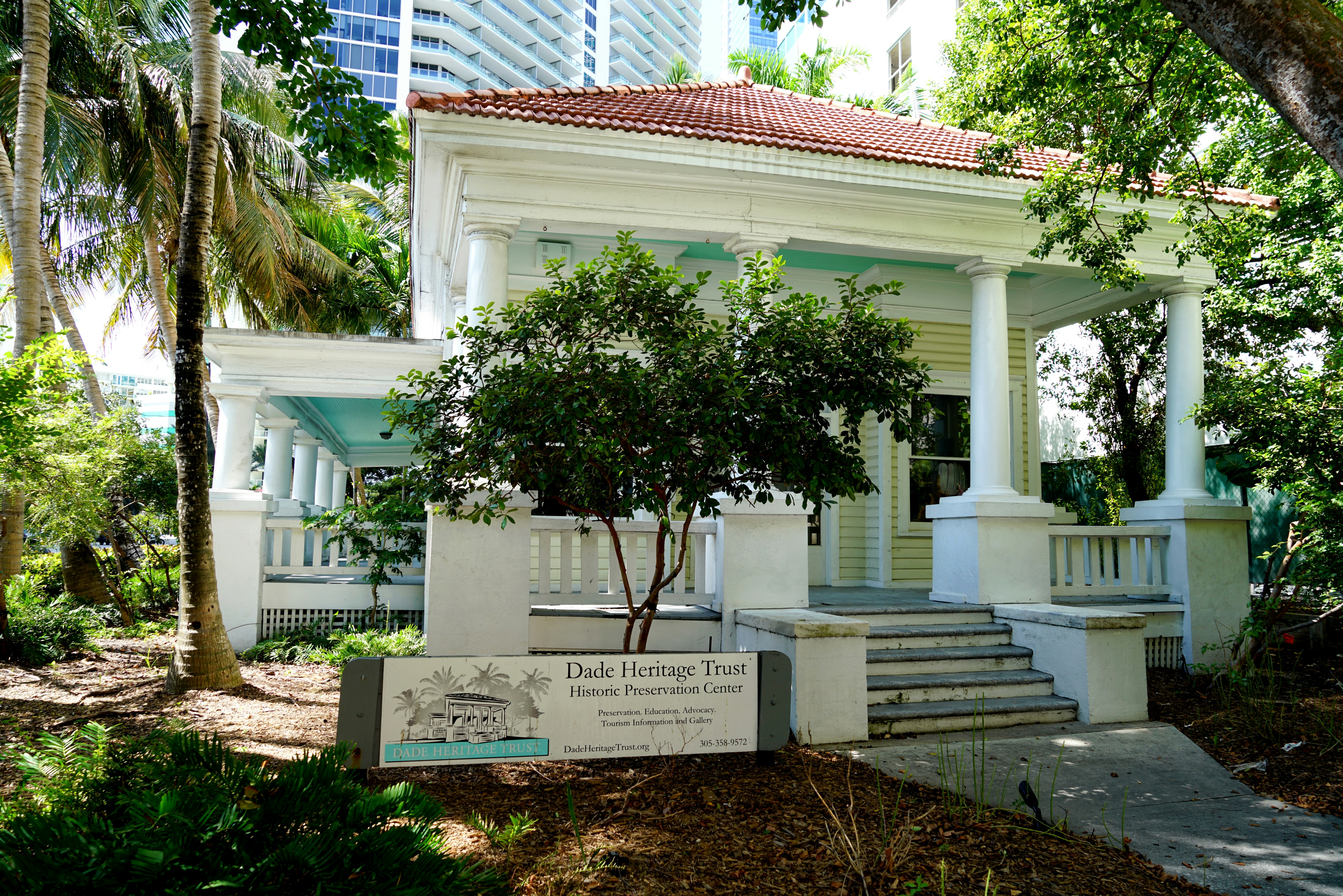 A small white heritage-style house with columns sits amid lush trees and a signboard in the foreground, conveying a tranquil courtyard scene.