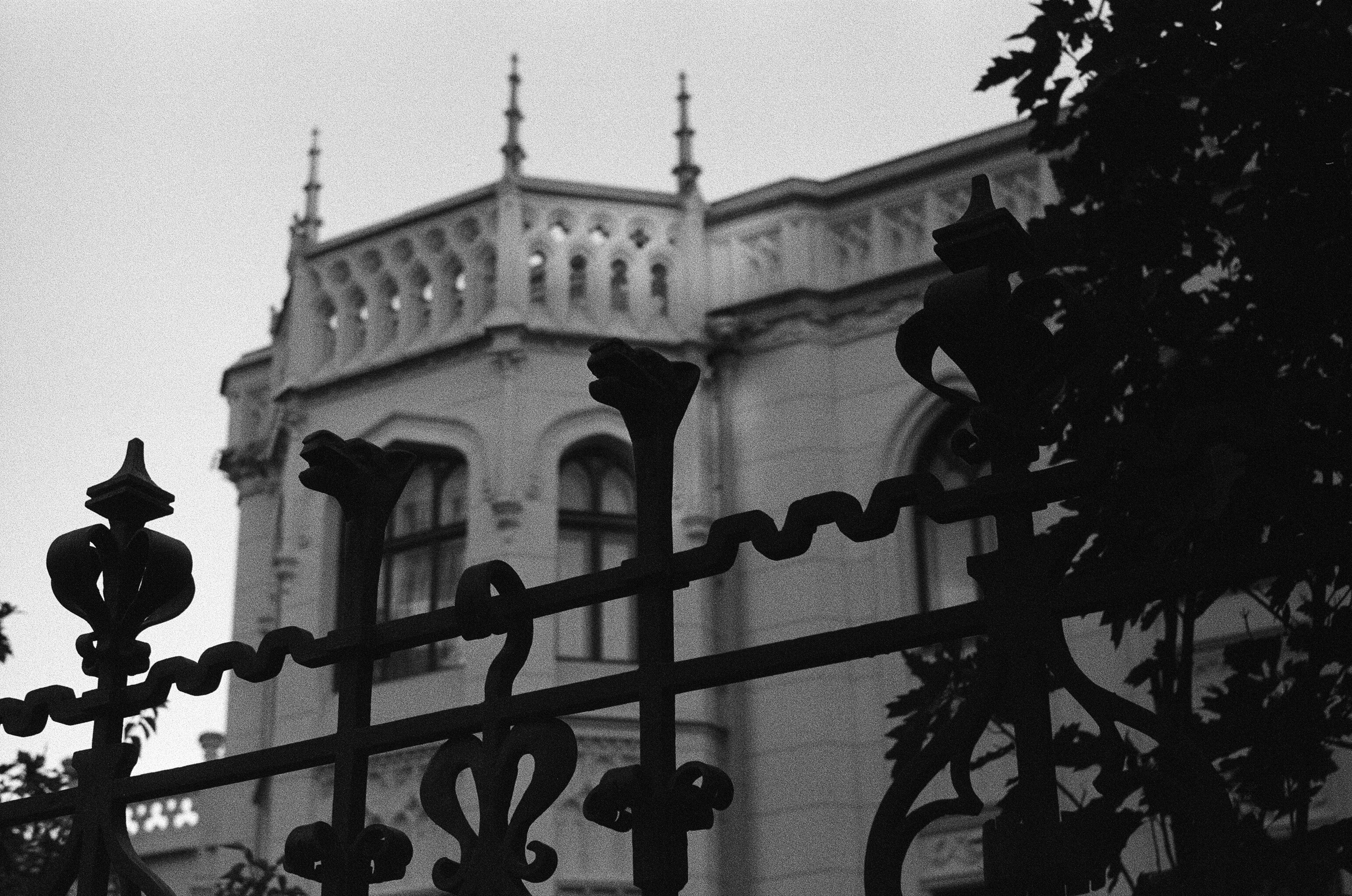 This dramatic black‑and‑white shot captures the Neo‑Gothic splendor of the Morozova Mansion on Spiridonovka Street in Moscow. A low‑angle view emphasizes the ornate balustrades, pointed arches, and decorative pinnacles. In the foreground, a wrought‑iron fence and tree silhouette frame the historic facade, creating a moody, textured composition.
