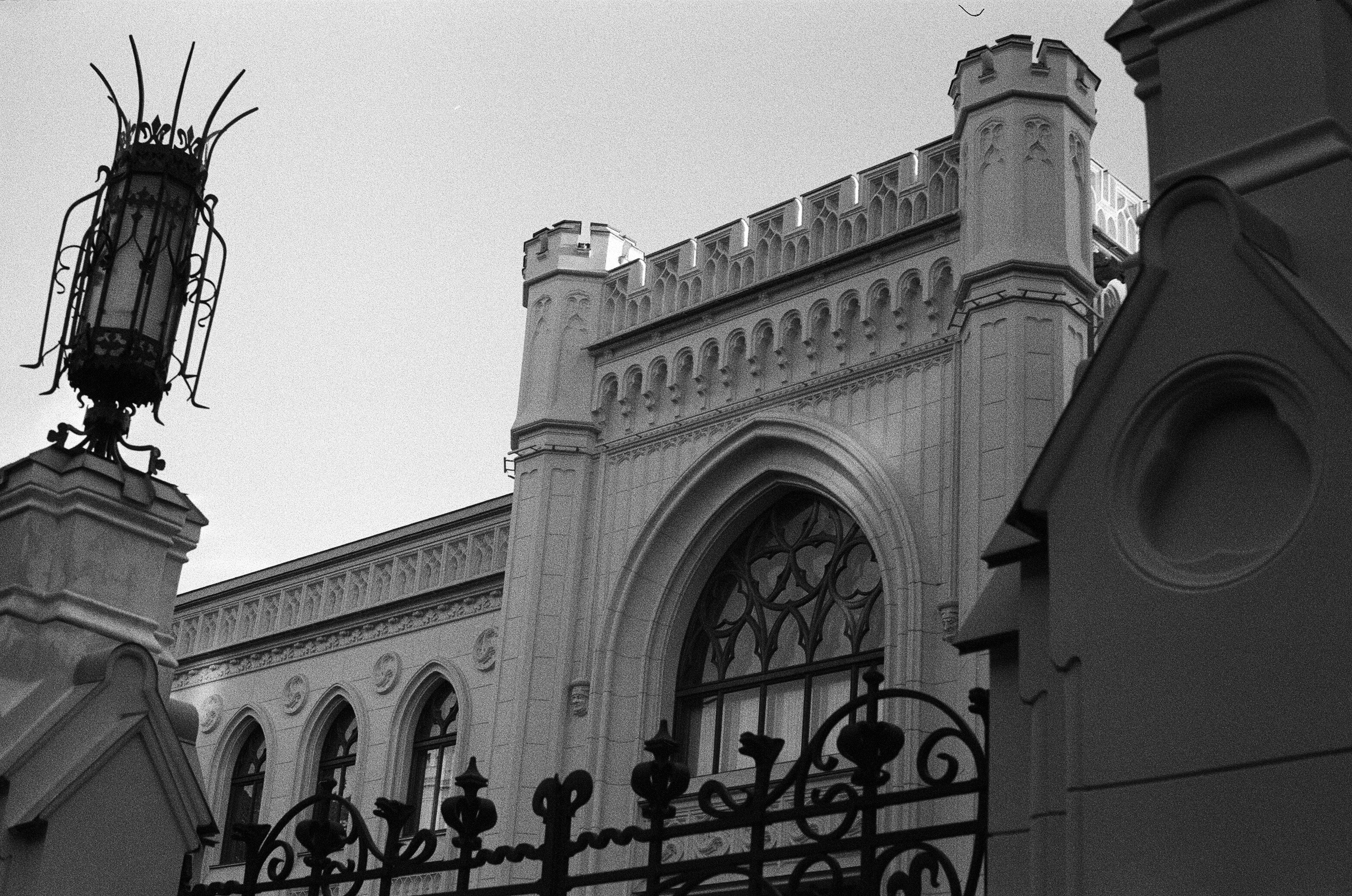 Monochrome architectural detail featuring ornate iron fencing, a prominent arched window, and decorative masonry on a grand gatehouse facade.