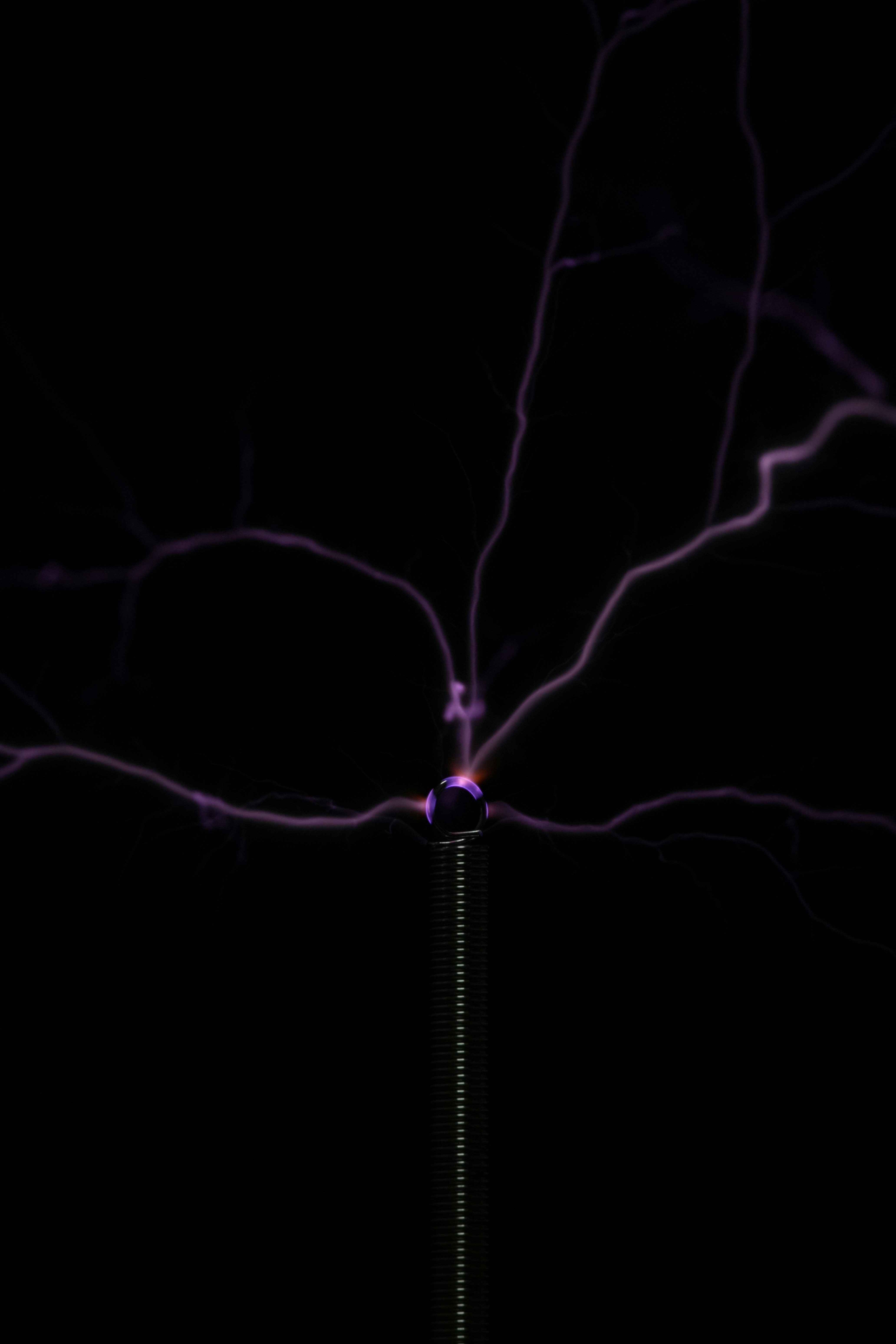 A single arc of purple lightning crowns a slender, ribbed metal rod set against a black background, emphasizing a center-aligned focus on the glowing crown and the top of the rod. This is a photographic study of electrical discharge and minimalistic composition.