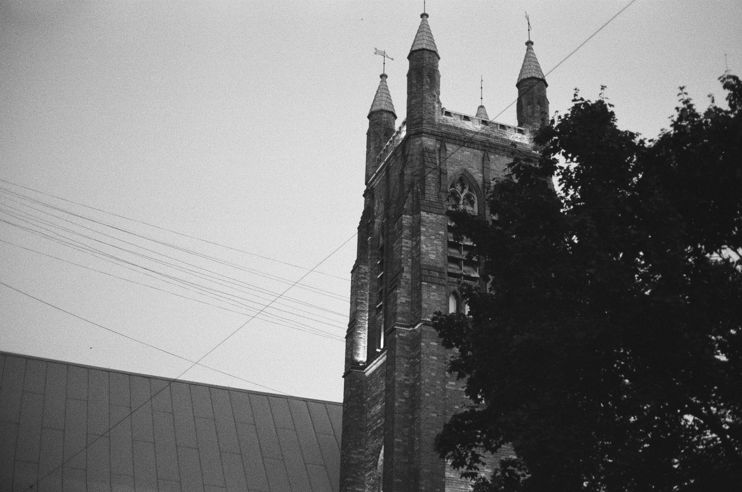 This moody black‑and‑white shot features the soaring tower of the Anglican Cathedral of St. Andrew in Moscow, rendered in Gothic Revival style. Pointed spires and lancet windows punctuate the textured stone facade, while tree silhouettes and power lines frame the composition, underscoring the building’s verticality and historic gravitas.