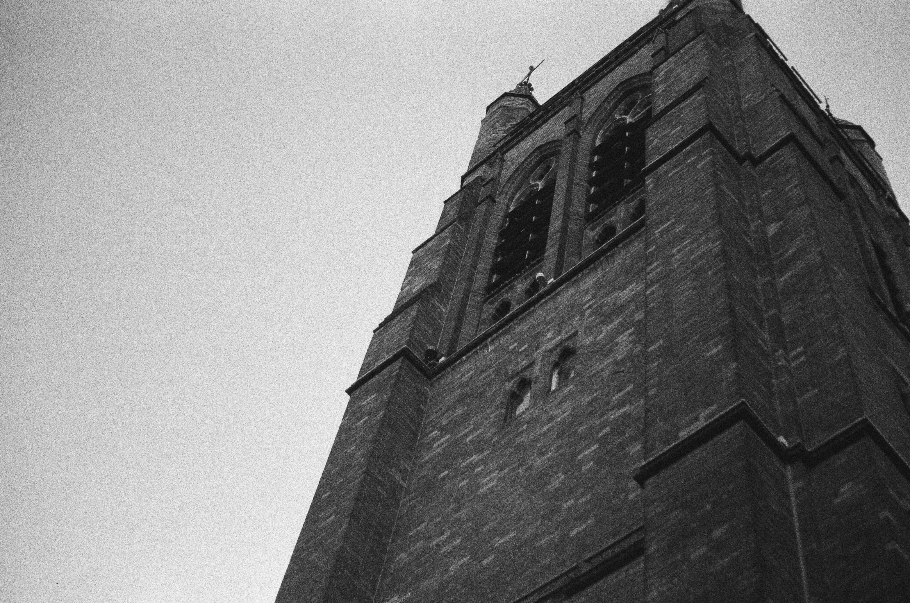 A towering stone church steeple dominates the frame, captured from a low angle that emphasizes its vertical lines and looming presence. The grayscale texture highlights masonry detail and architectural depth.