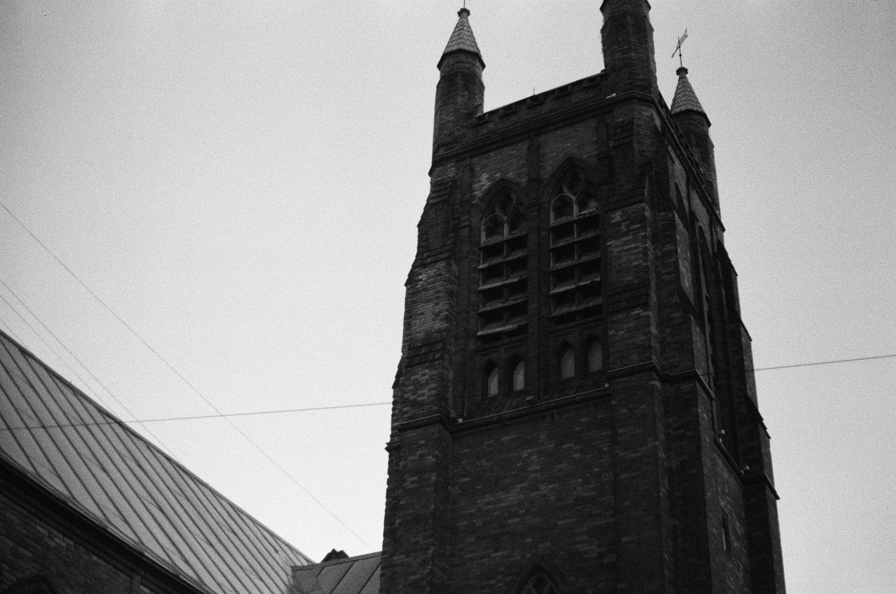 Monochrome view of a tall stone church tower rising over a sloped metal roof and power lines, emphasizing architectural texture and verticality. This is a photographic study of imposing vertical structure.